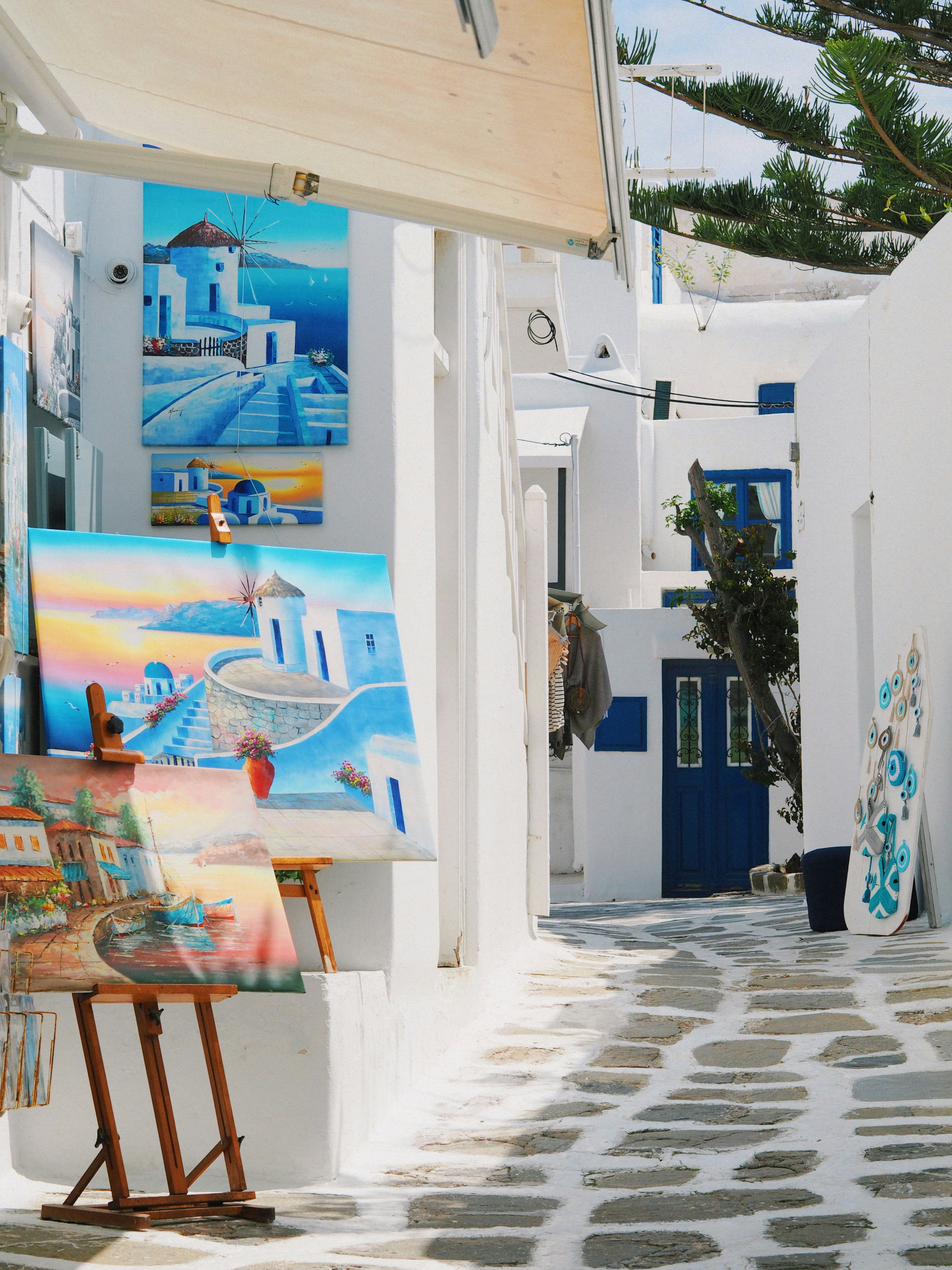 Colorful seascape paintings lean on easels along a sunlit white-washed alley, with cobalt doors and a stone-paved path.