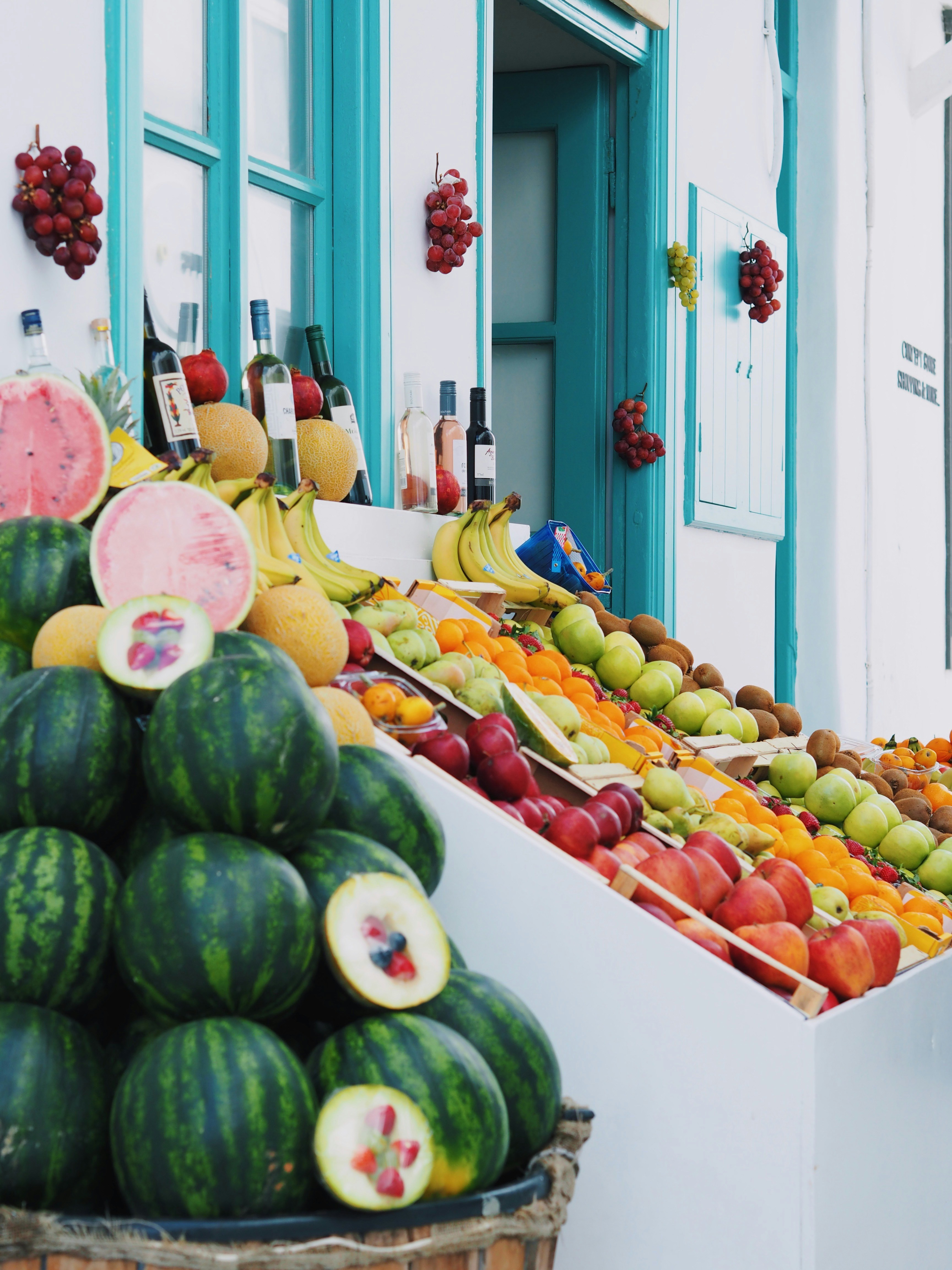 Vibrant fruit display cascades along a white Cycladic wall beneath turquoise doors, featuring stacked watermelons, citrus, and bottles. The arrangement blends market energy with bold color contrast.