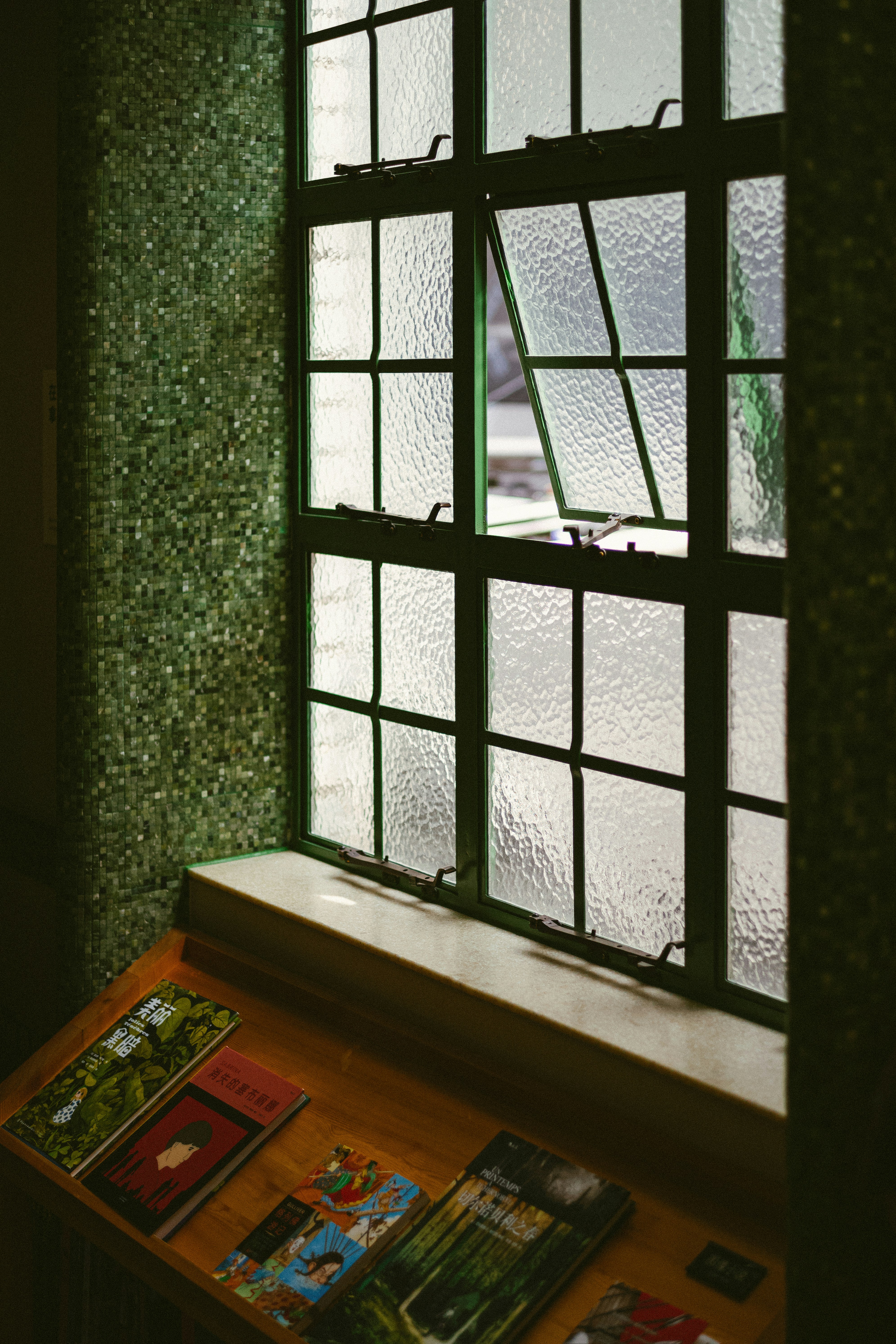 Frosted grid window set in a green tiled wall, with a wooden sill displaying colorful magazines.