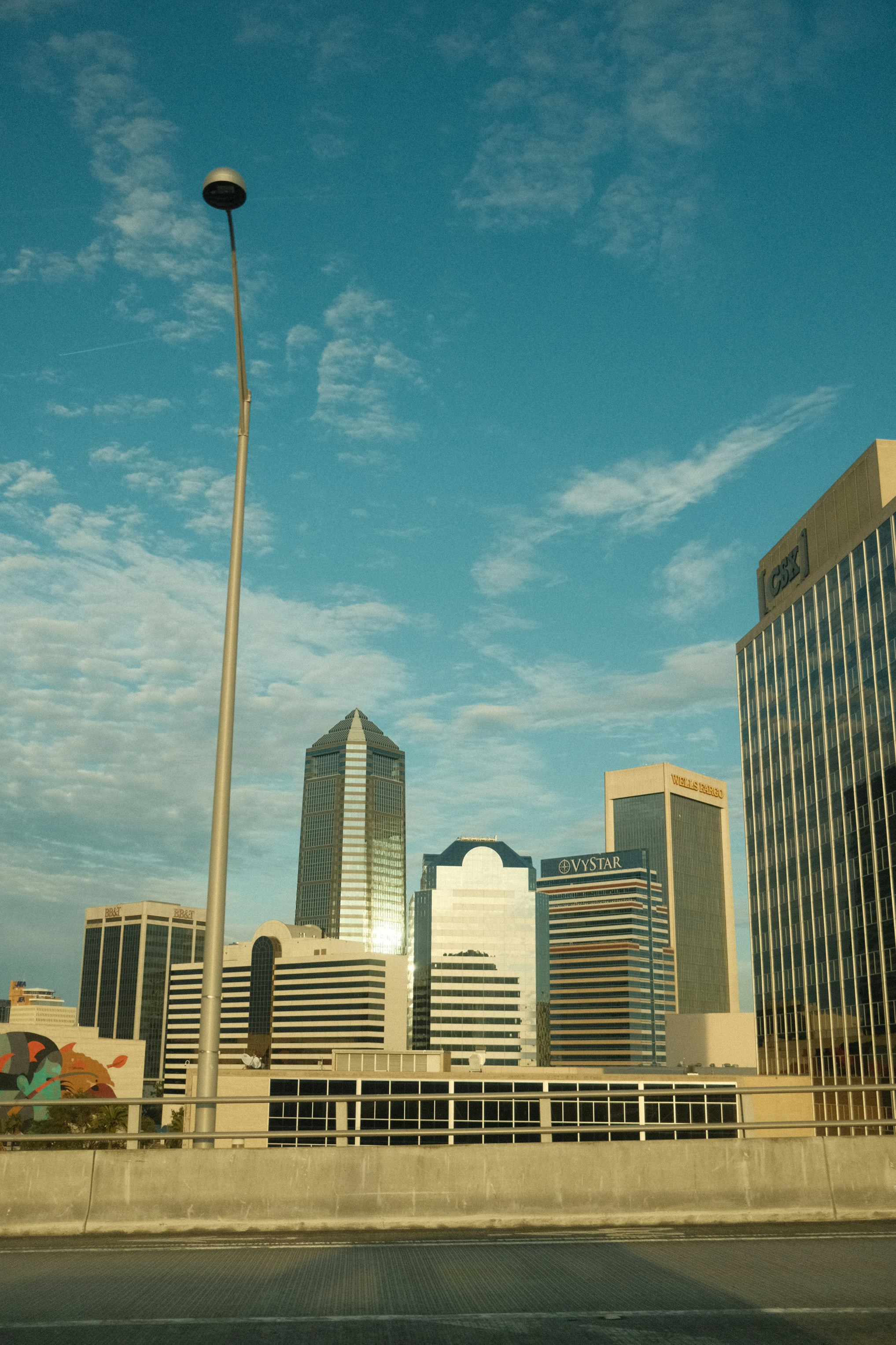 City skyline with towering buildings and a prominent streetlight under a blue sky with scattered clouds.