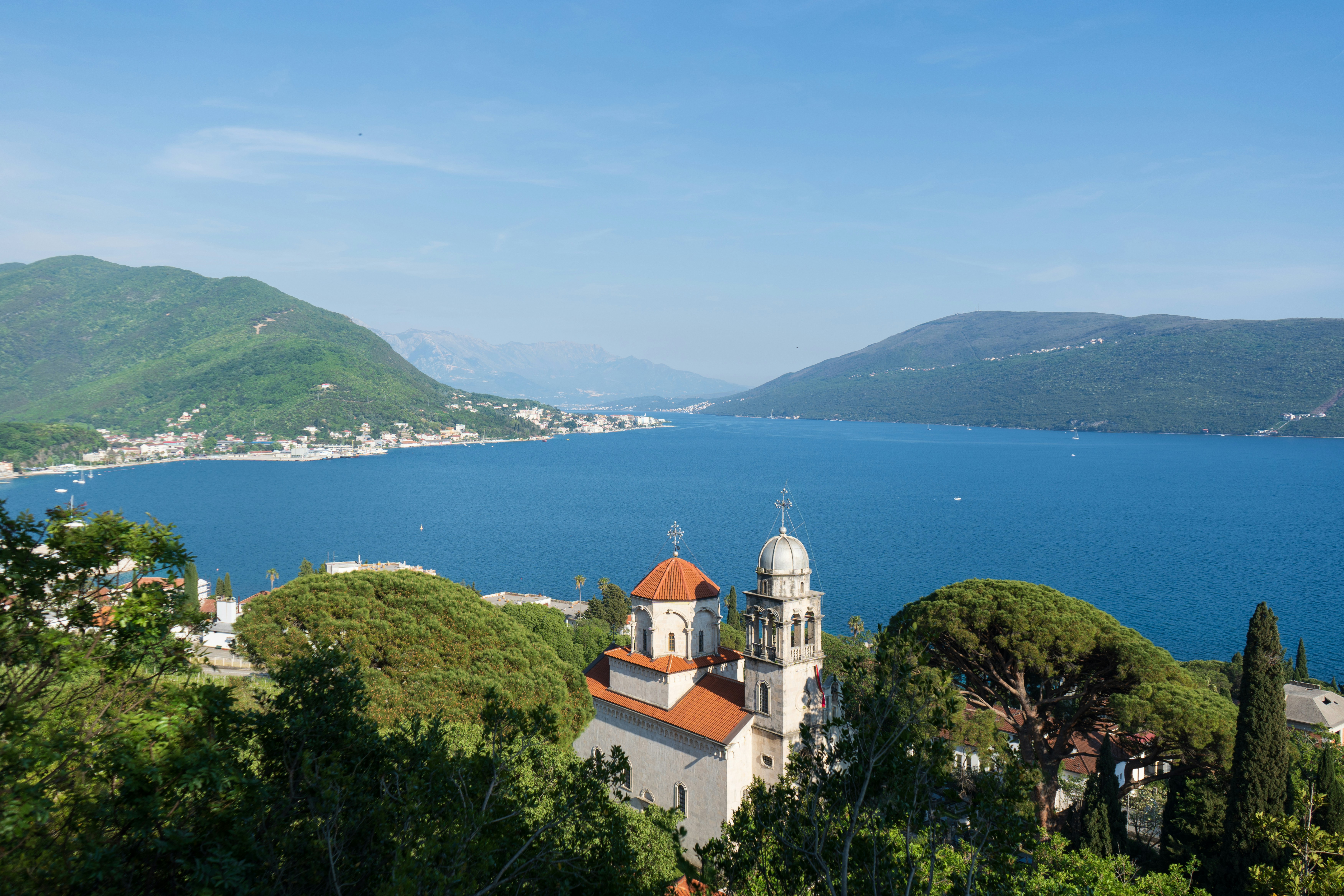 Church with red-tiled roof nestled among trees overlooking a vast blue bay and distant hills under a clear sky.