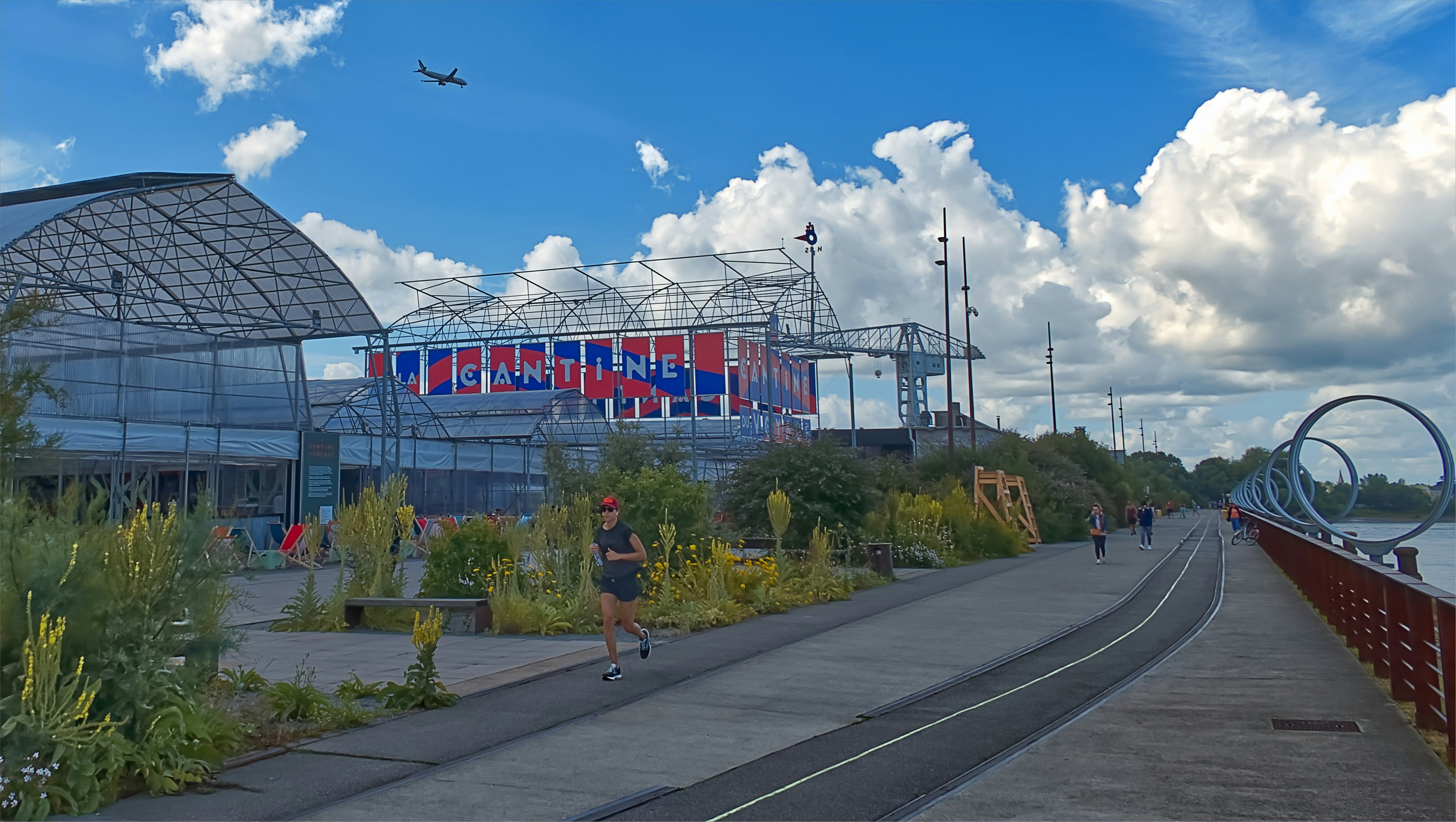 Jogger on a sunlit riverside path beside a glass-roof pavilion under a plane-filled sky