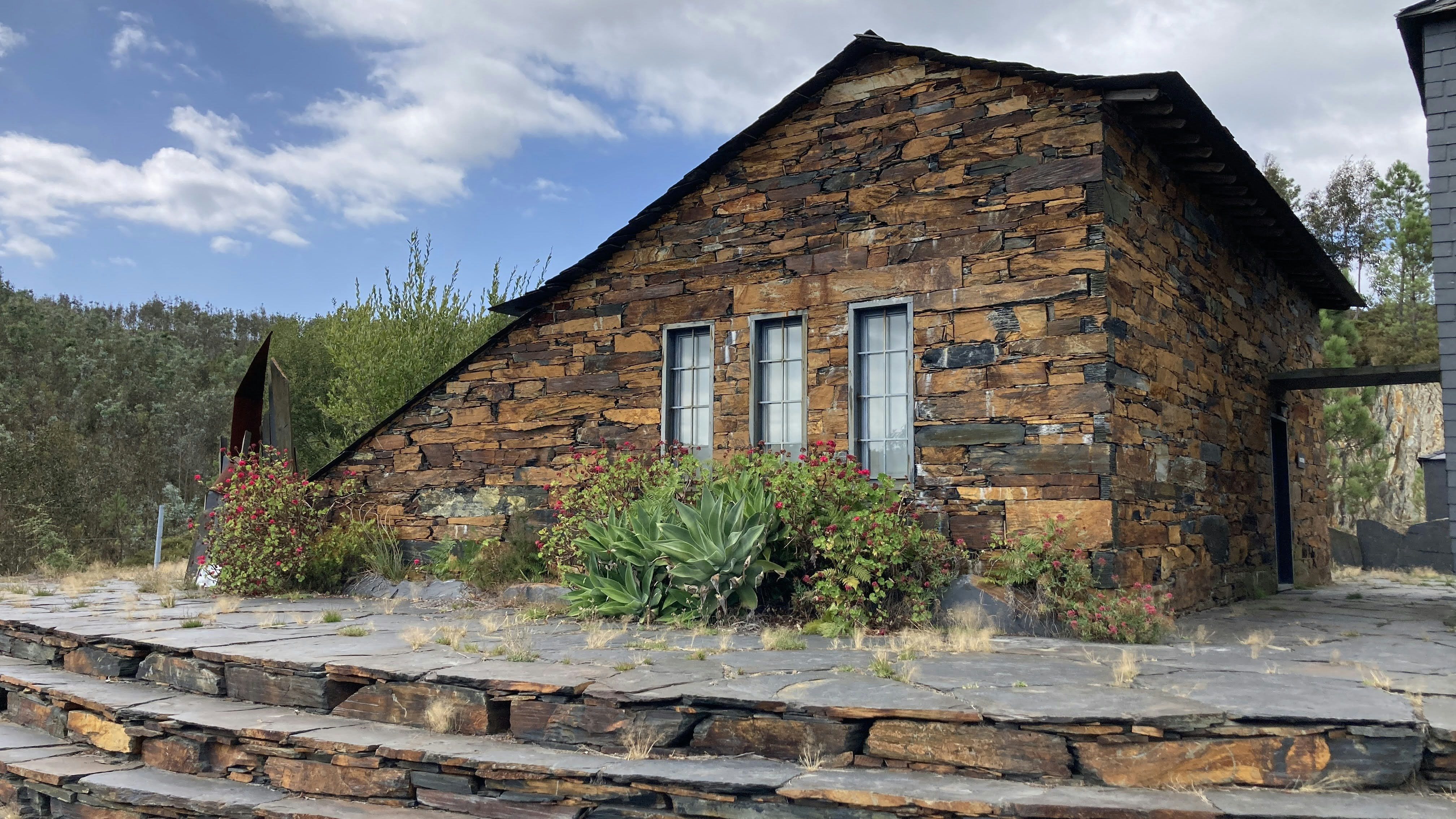 A stone cabin with weathered walls surrounded by greenery, set against a partly cloudy sky.