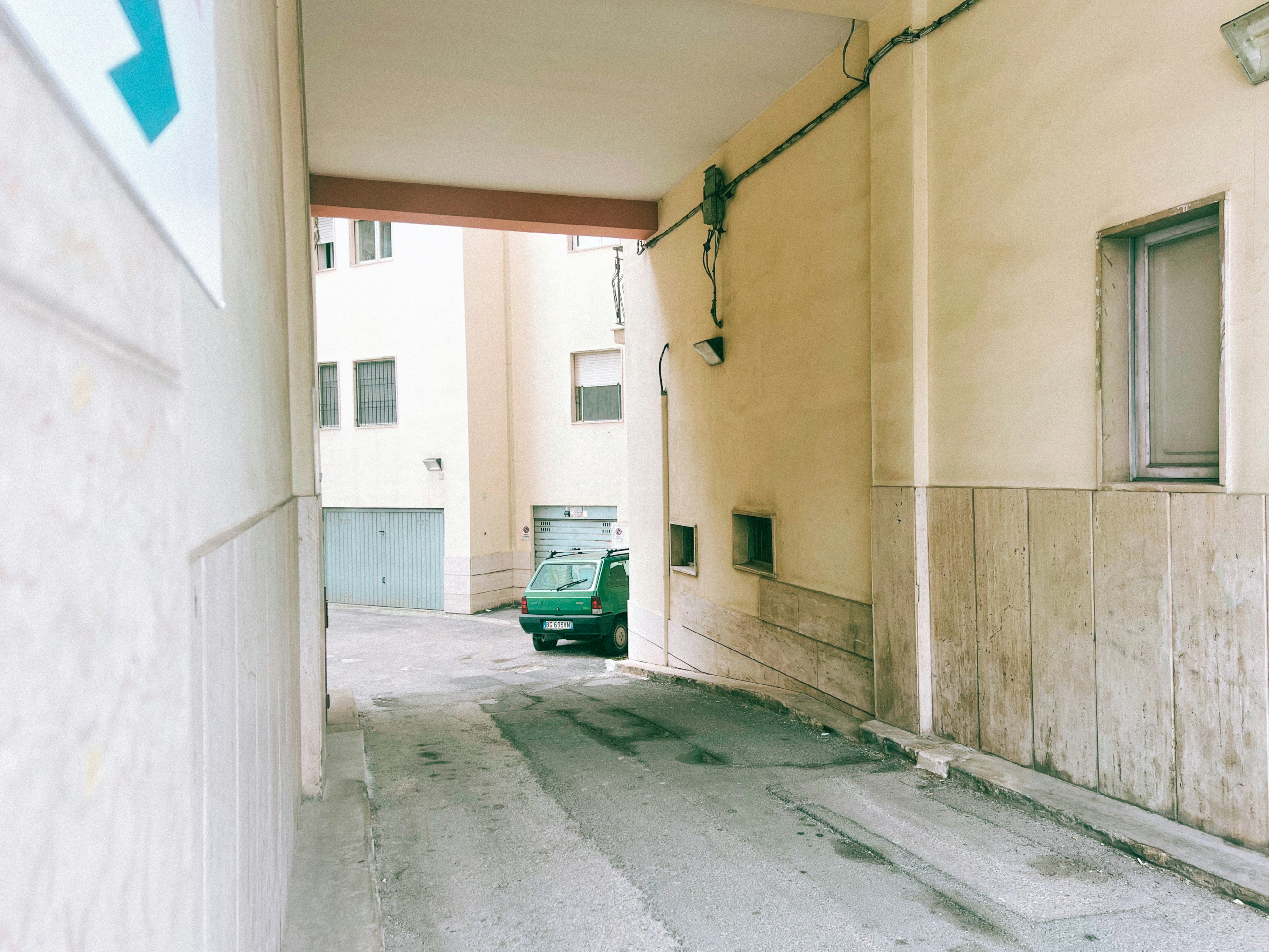 Narrow alleyway with a green vehicle parked at the end, framed by beige building walls and overhead beams.