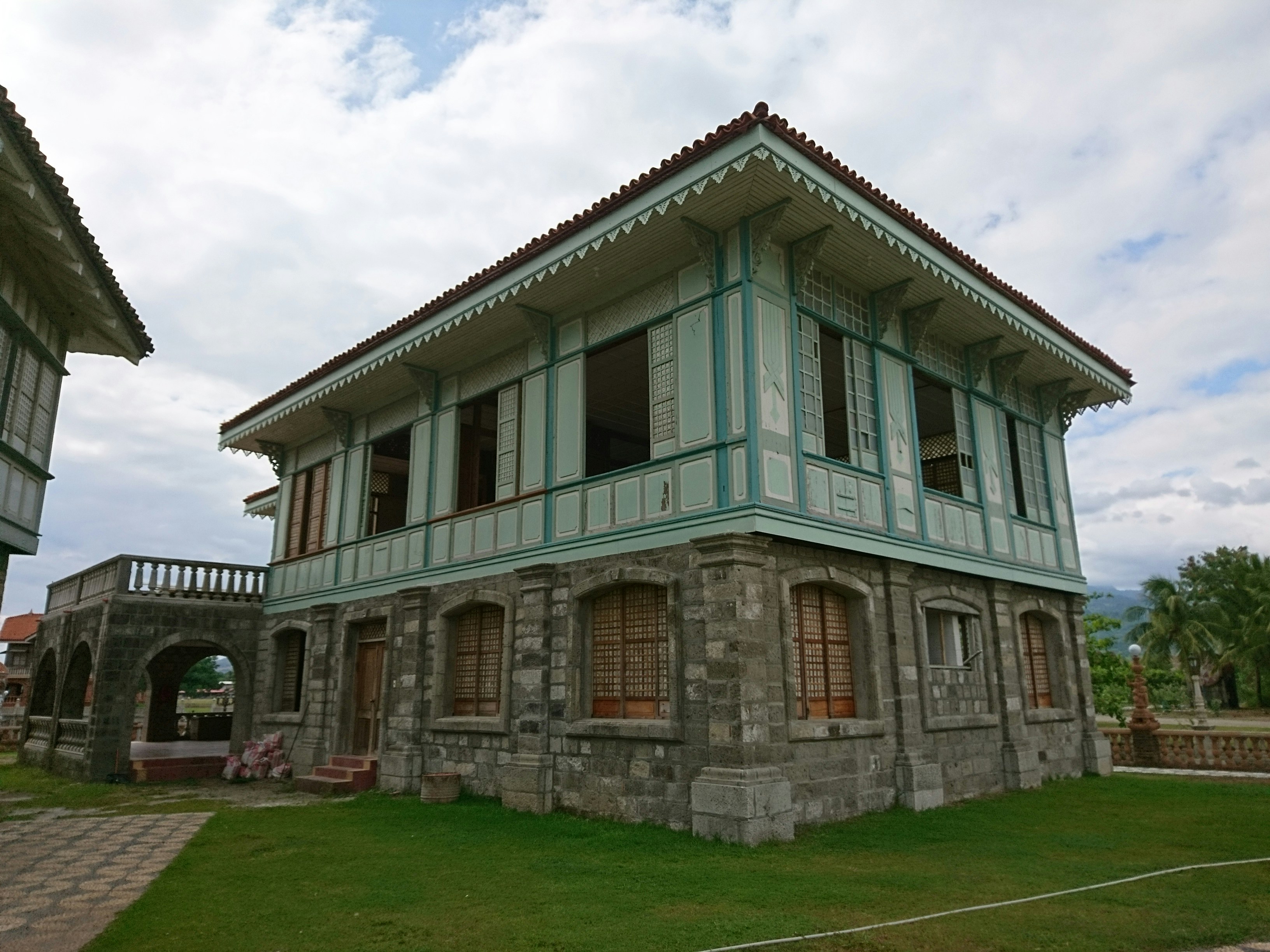 Two-story villa with a stone base and mint-green upper level sits on a manicured lawn beneath a cloudy sky. Architectural details emphasize colonial charm.