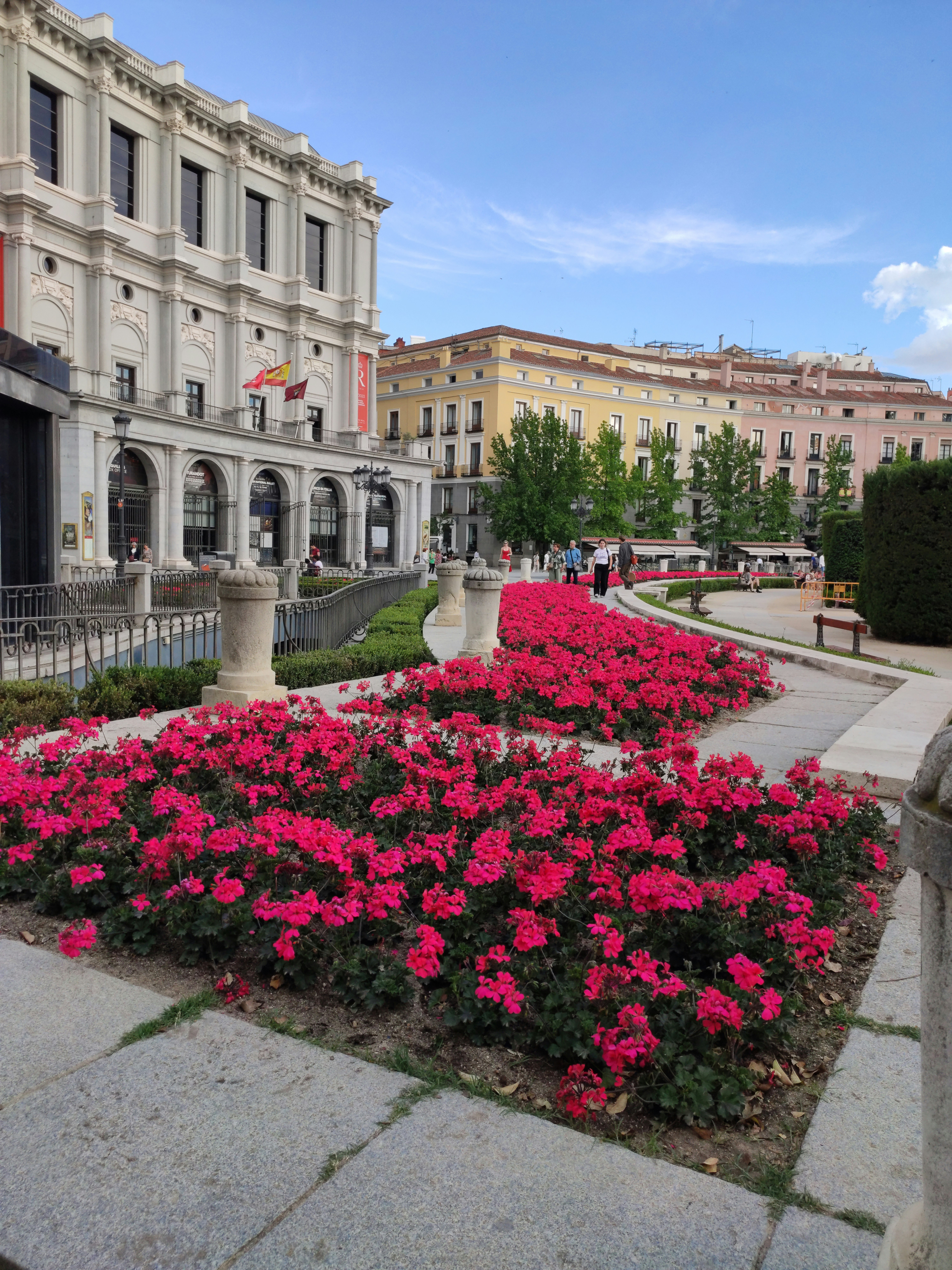 Vibrant pink flower beds line a sunlit plaza with historic architecture and a stone balustrade along a water feature.