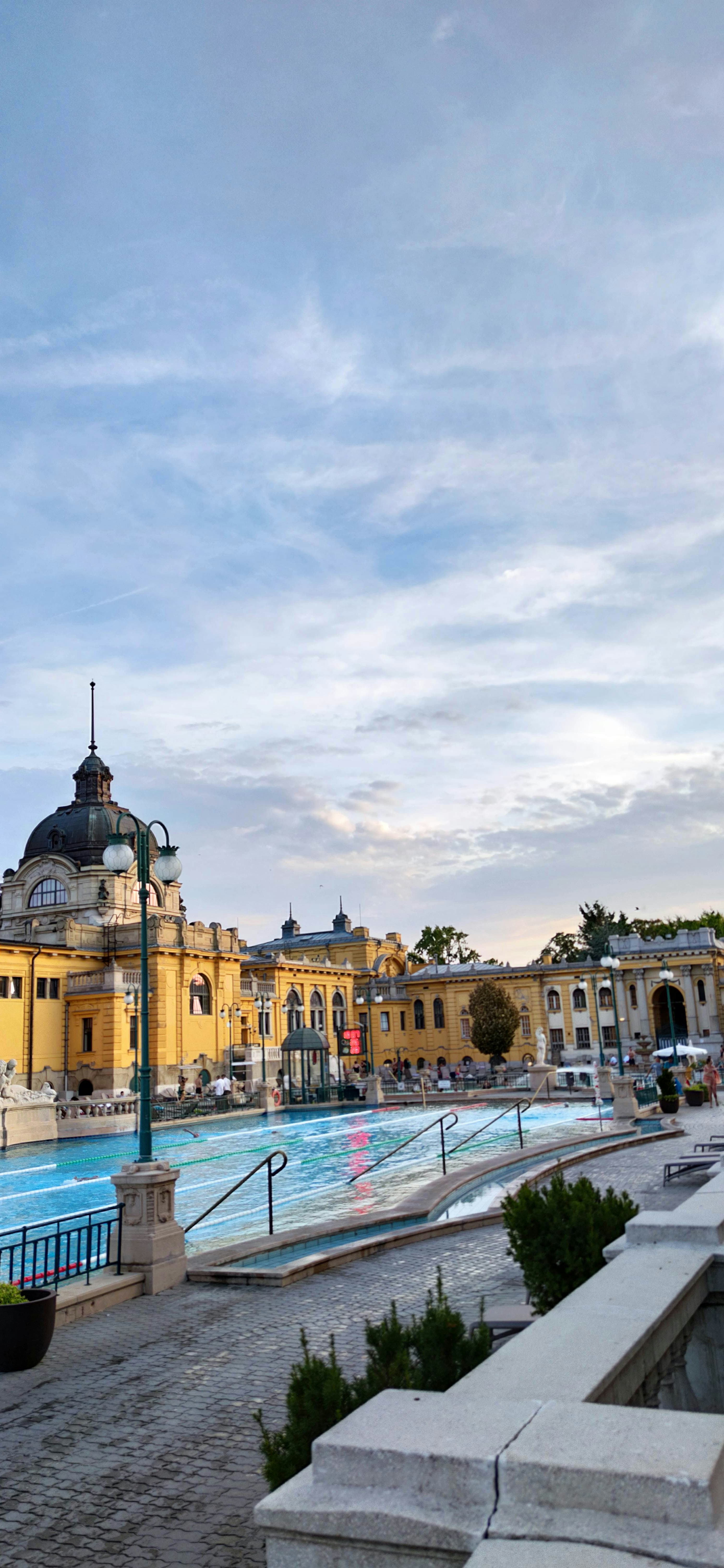 Photograph of the Széchenyi Thermal Bath complex in Budapest, featuring a bright blue pool and yellow domed buildings under a soft, expansive sky.