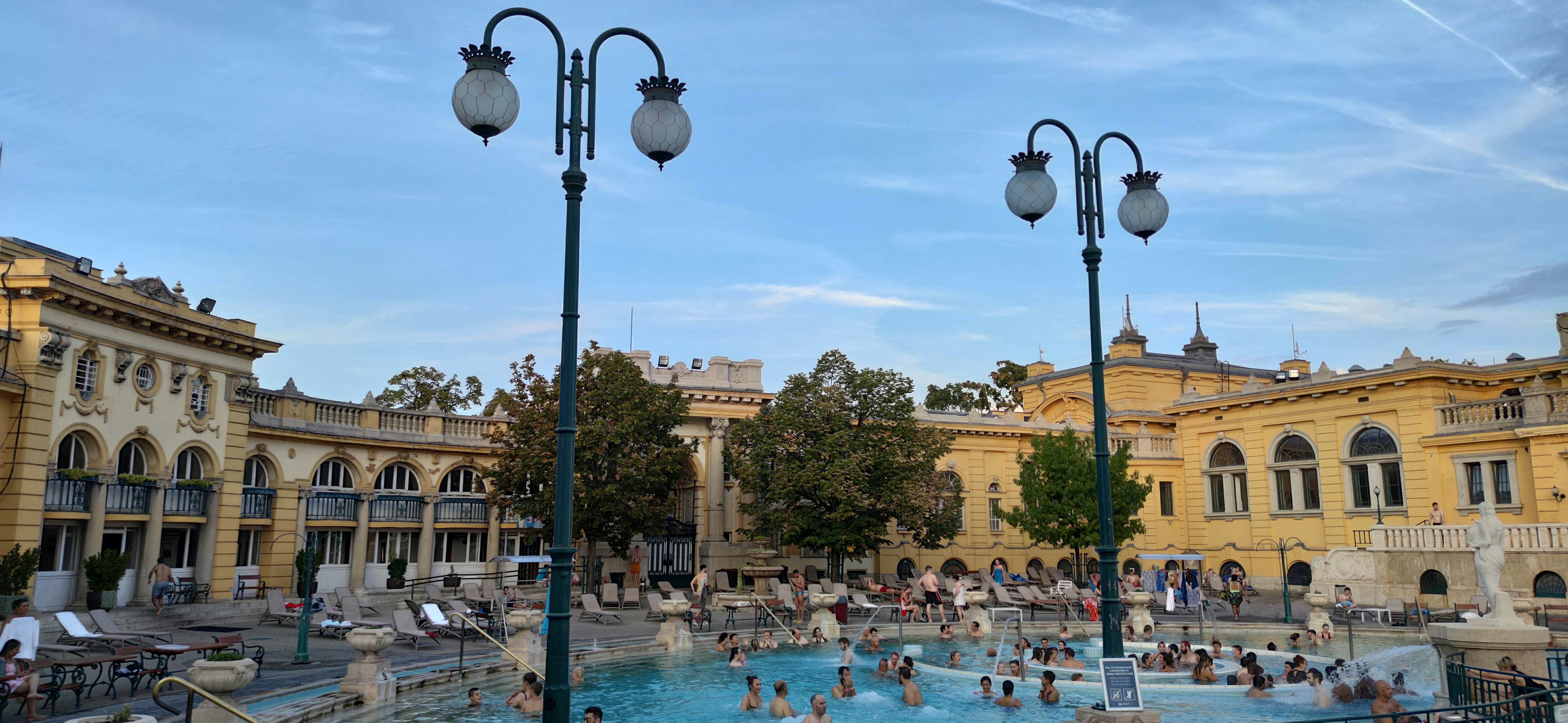 Outdoor pool scene framed by yellow historic façades and vintage lamp posts. Bathers relax along the blue water.