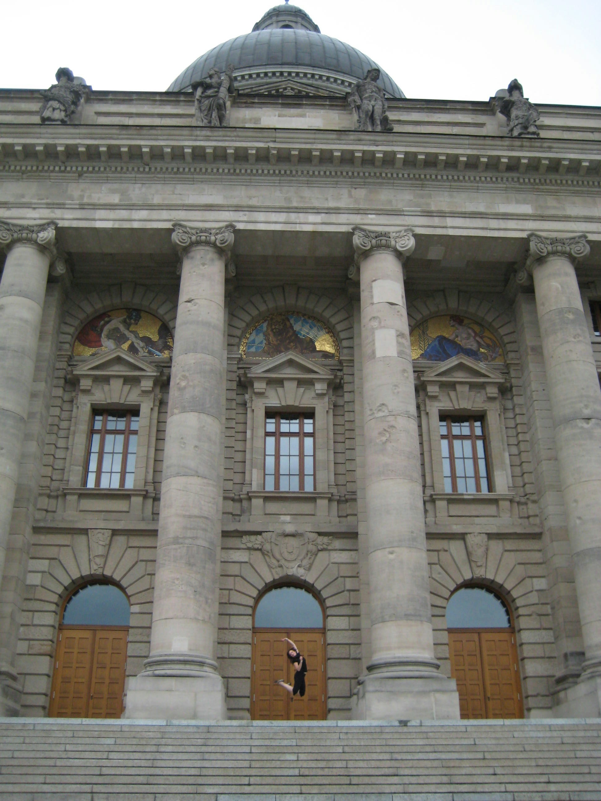 A lone dancer stands on the broad staircase in front of a neoclassical building with tall columns and a central dome. The scene emphasizes architectural grandeur and the dancer's poised silhouette against the monumental facade.