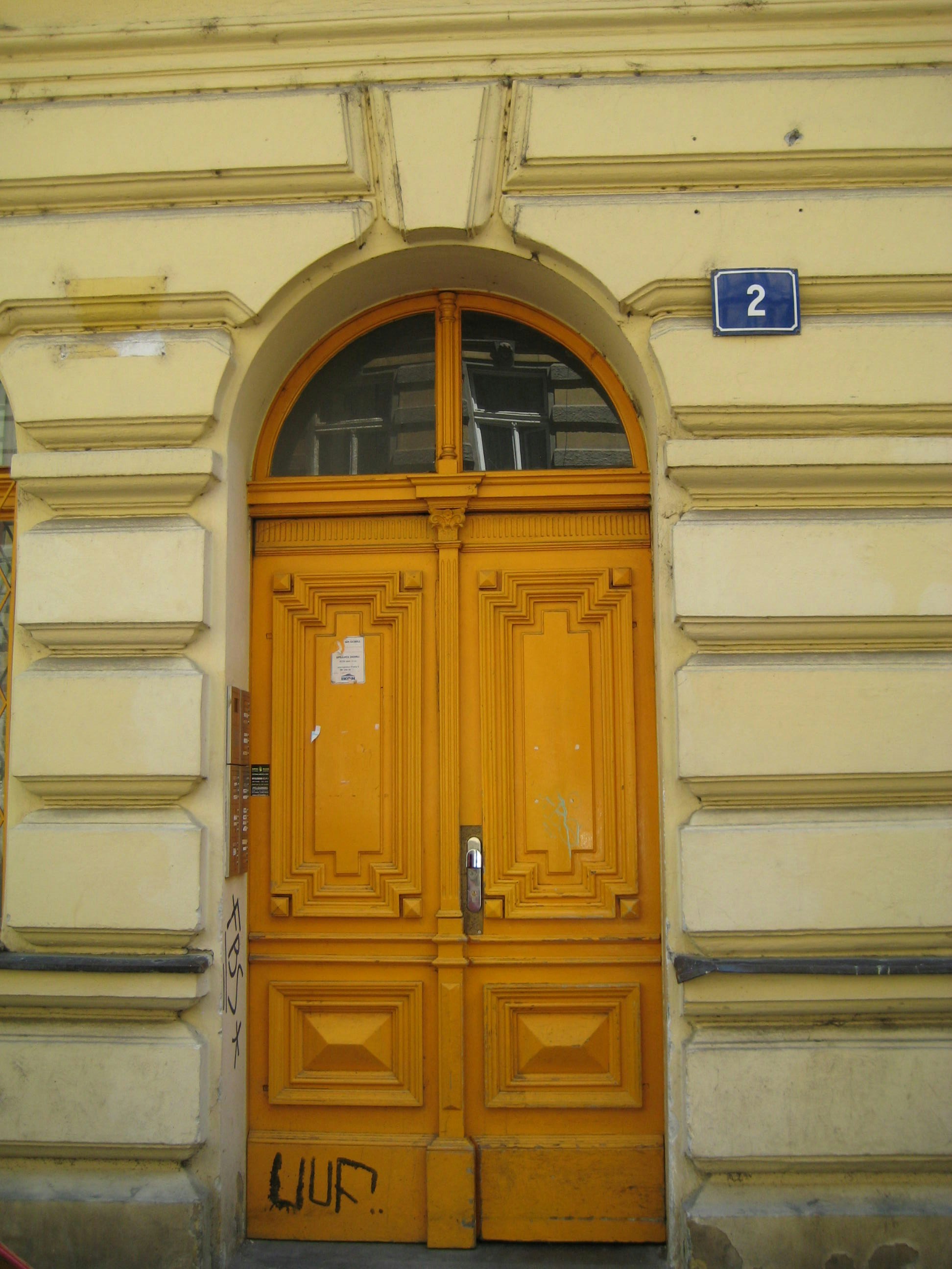 An amber, ornately panelled door framed by a cream neoclassical facade beneath an arched window. Urban details include a blue '2' plaque and subtle graffiti at the lower panel.