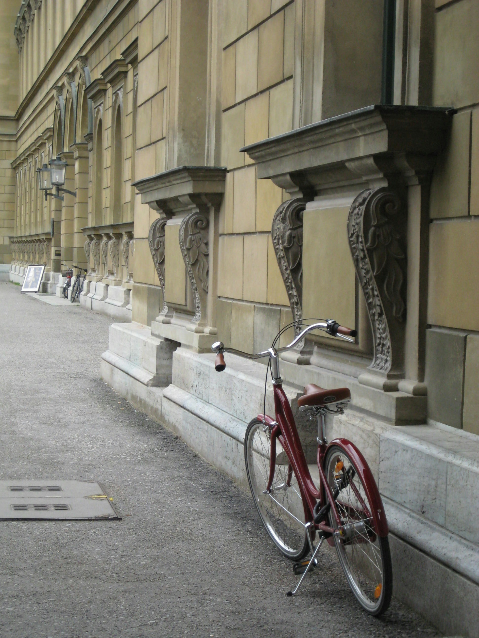 Photograph of a red bicycle leaning against a carved stone wall with ornate corbels along a sidewalk. The scene emphasizes architectural details and quiet urban charm.