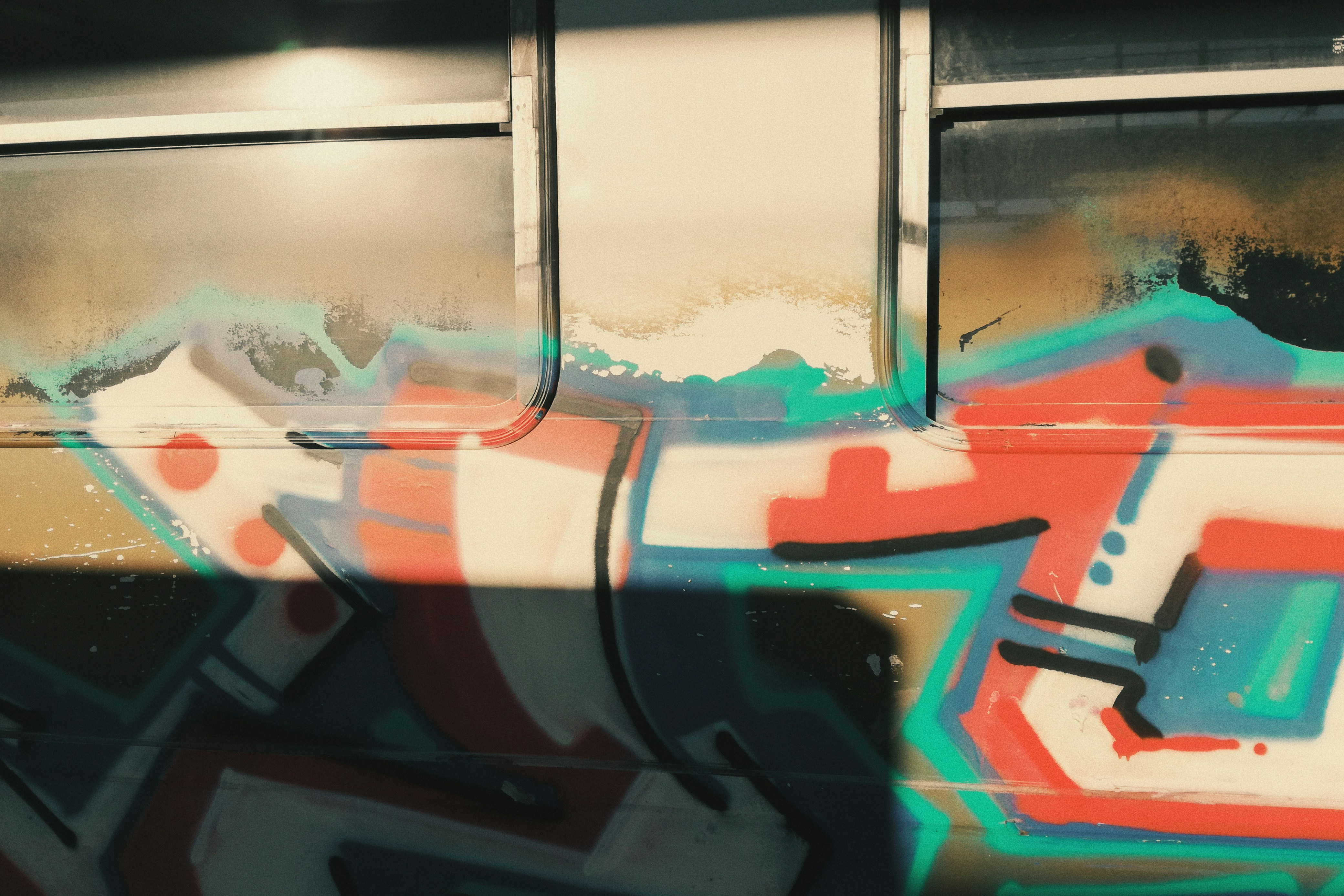Vibrant graffiti painting spans the lower panels of a train carriage, with teal, red, and orange shapes weaving across ribbed metal. Bright daylight highlights the bold color blocks and crisp lines of the mural.