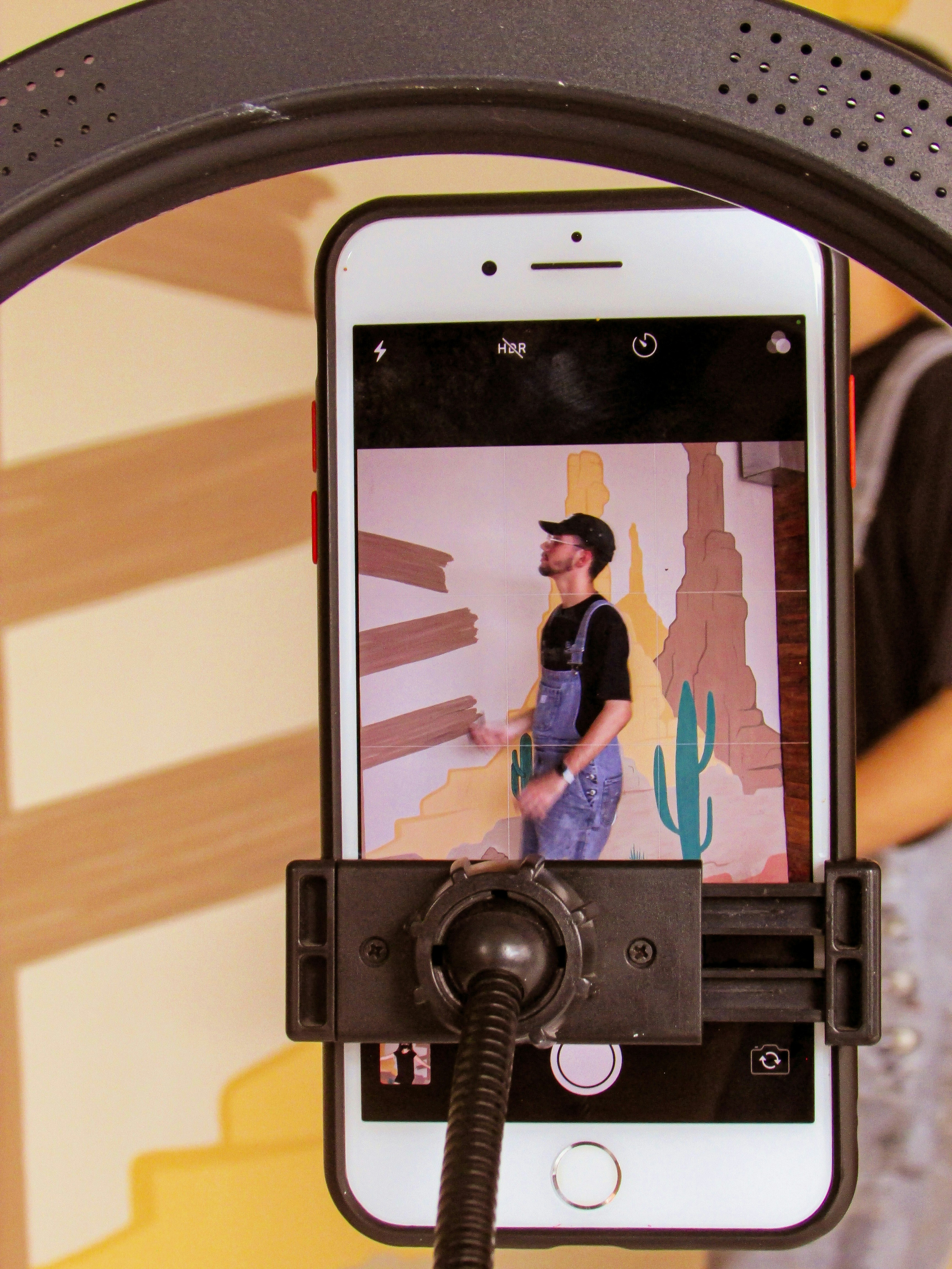 Phone screen reveals a painted desert backdrop with a bearded man in overalls, captured within a ring-light rig.