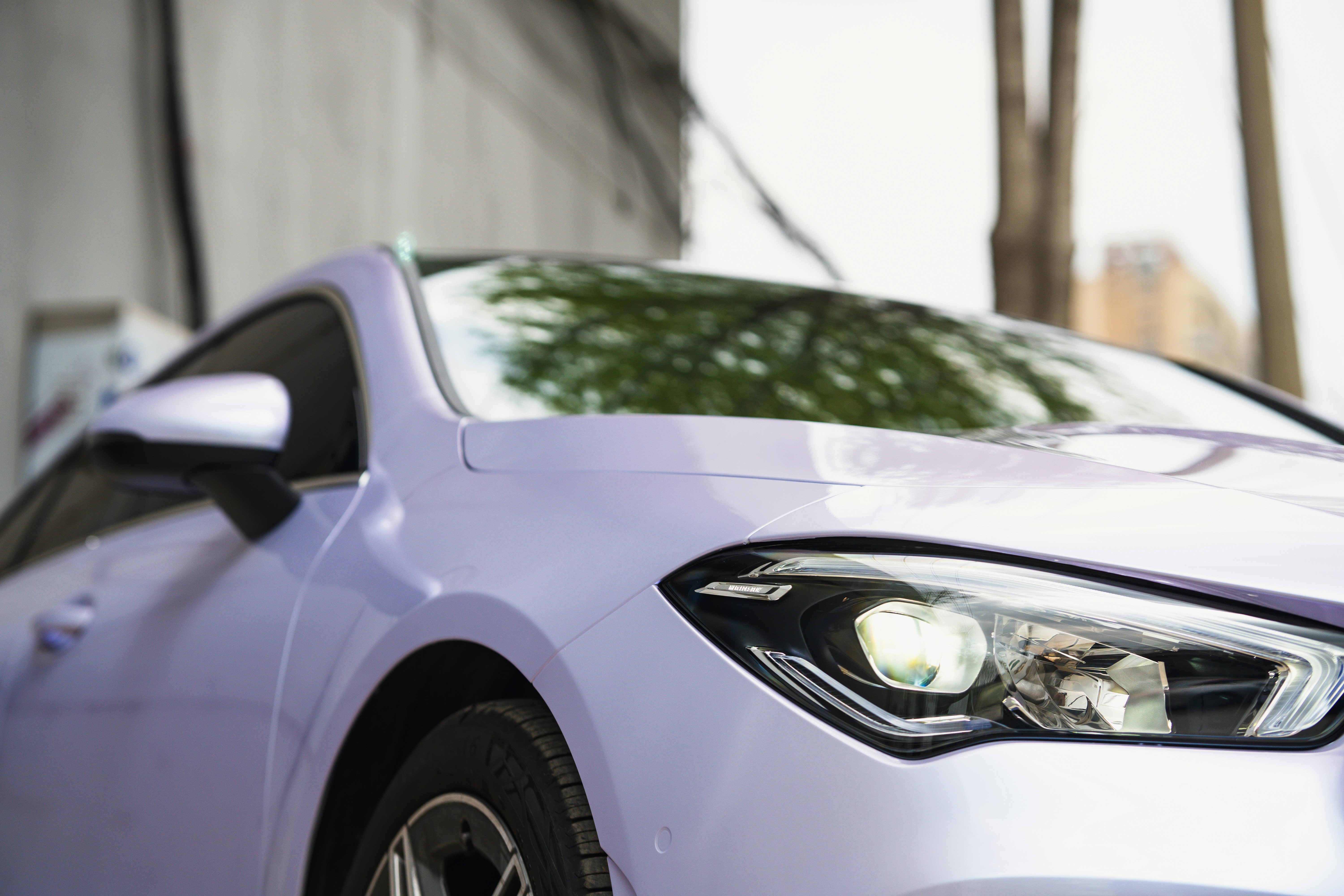Lilac Coupe Close-Up: Glowing Headlight and Curved Body Under Urban Dappled Light