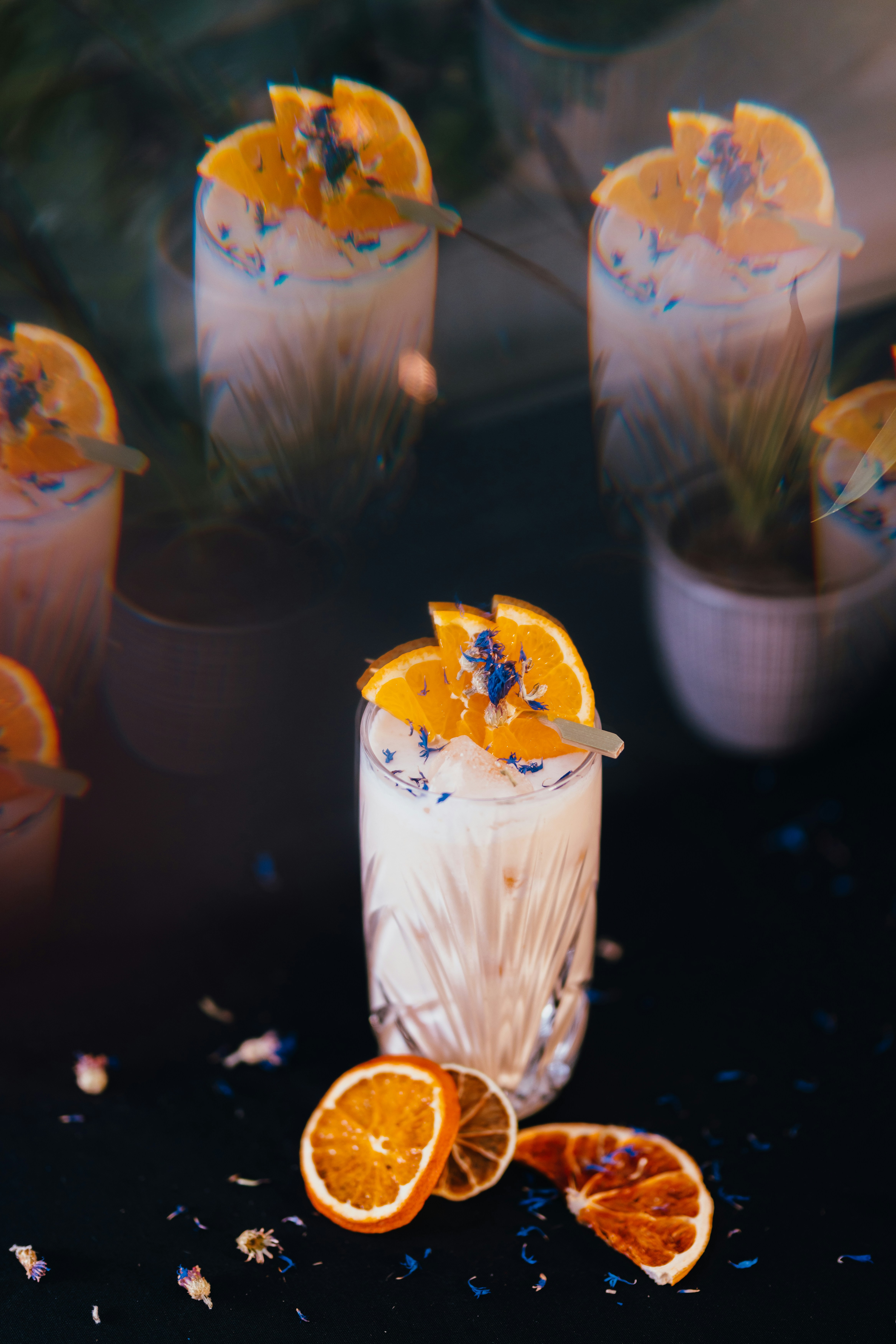 Tall glass with orange slices and lavender, surrounded by reflected images on dark surface.