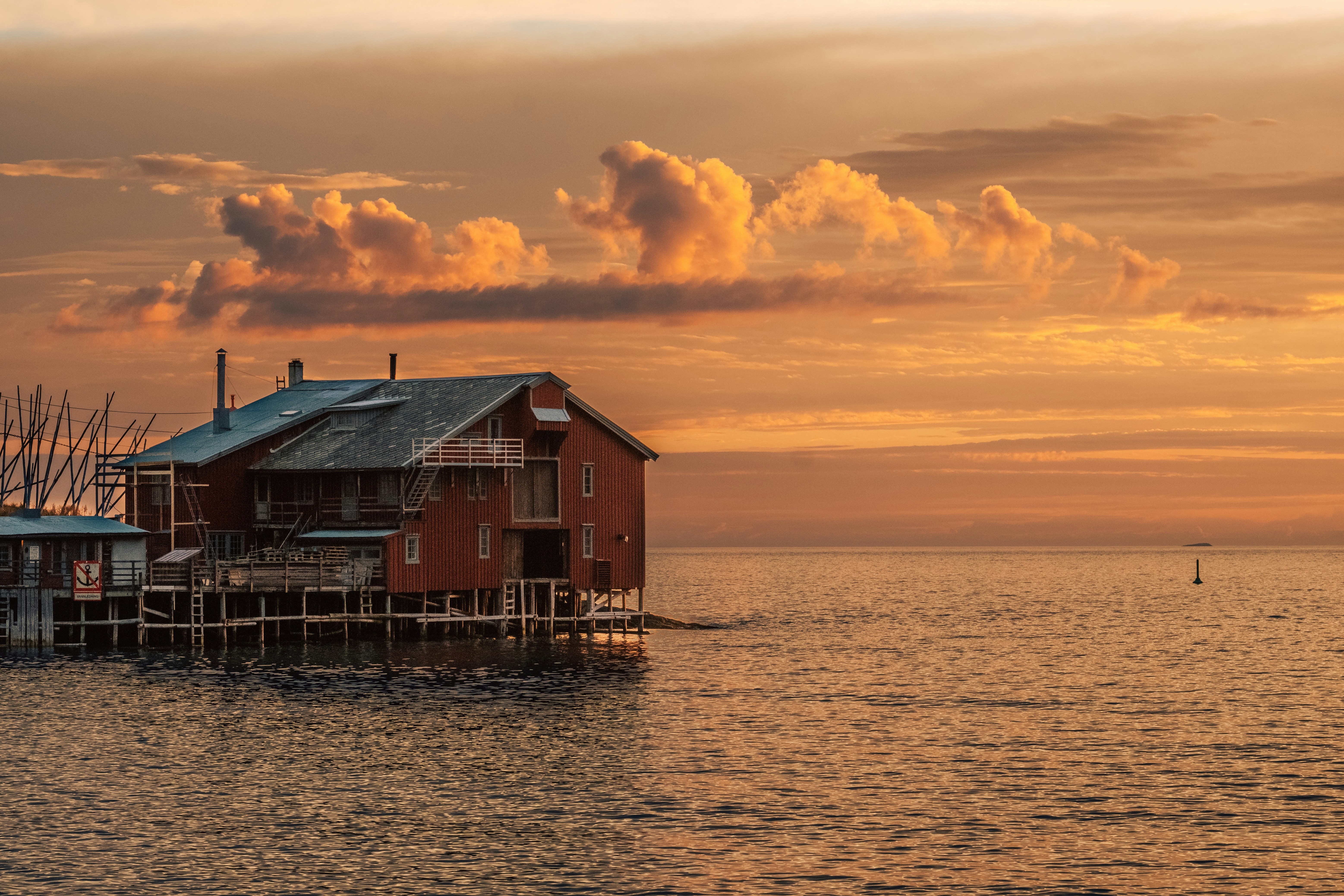 Red fishing house on stilts over calm water at sunset with dramatic clouds illuminating the sky.