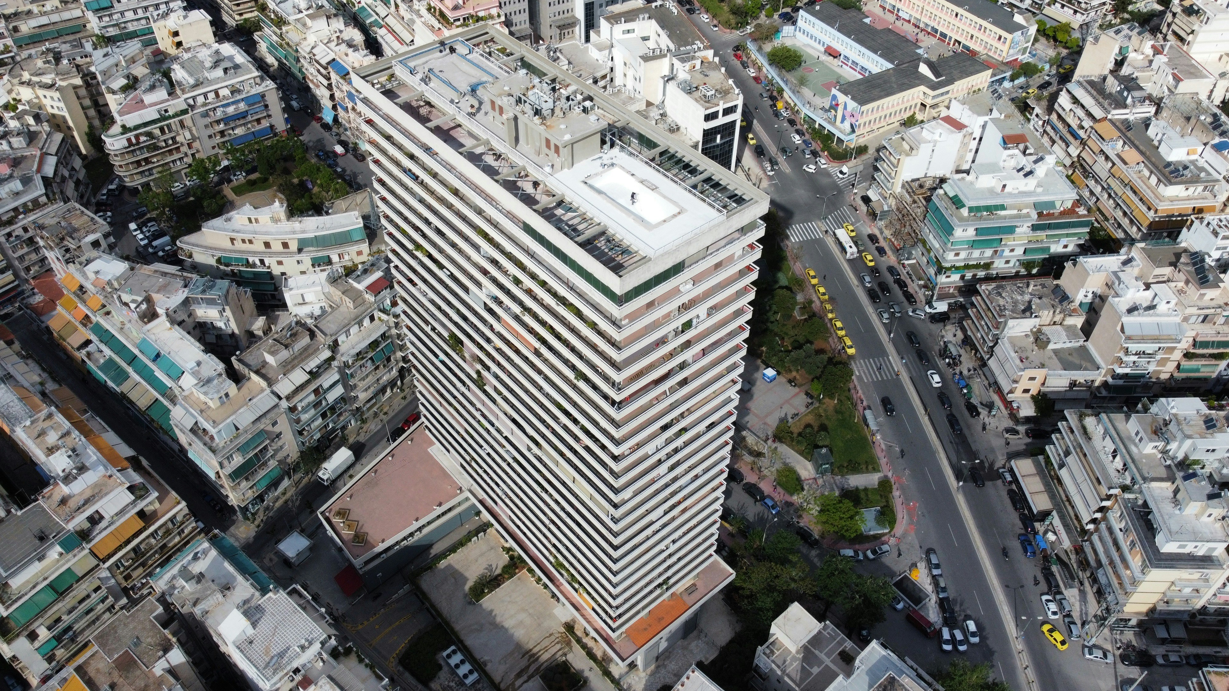 This aerial image captures a towering skyscraper amidst a bustling urban landscape, showcasing the intricate layout of streets and buildings. The composition highlights the stark contrast between the modern architecture of the skyscraper and the surrounding traditional structures. The image is bathed in natural light, enhancing the muted earth tones of the buildings against the vibrant greenery and the dynamic pattern of streets below, creating a visually striking cityscape.