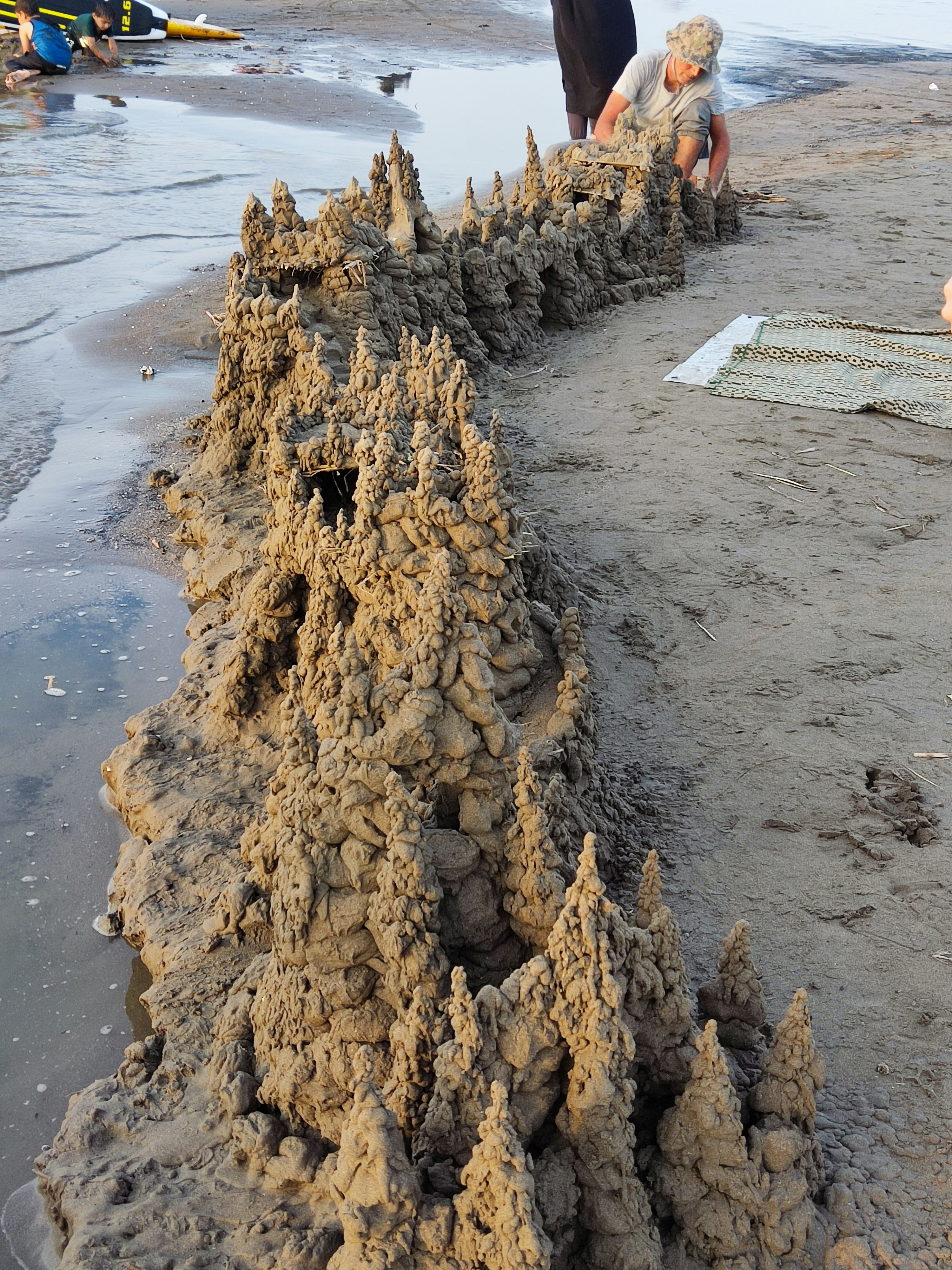 Intricate sandcastle resembling a mountain range, crafted by beachgoers, with individuals engaged in the creative process nearby.