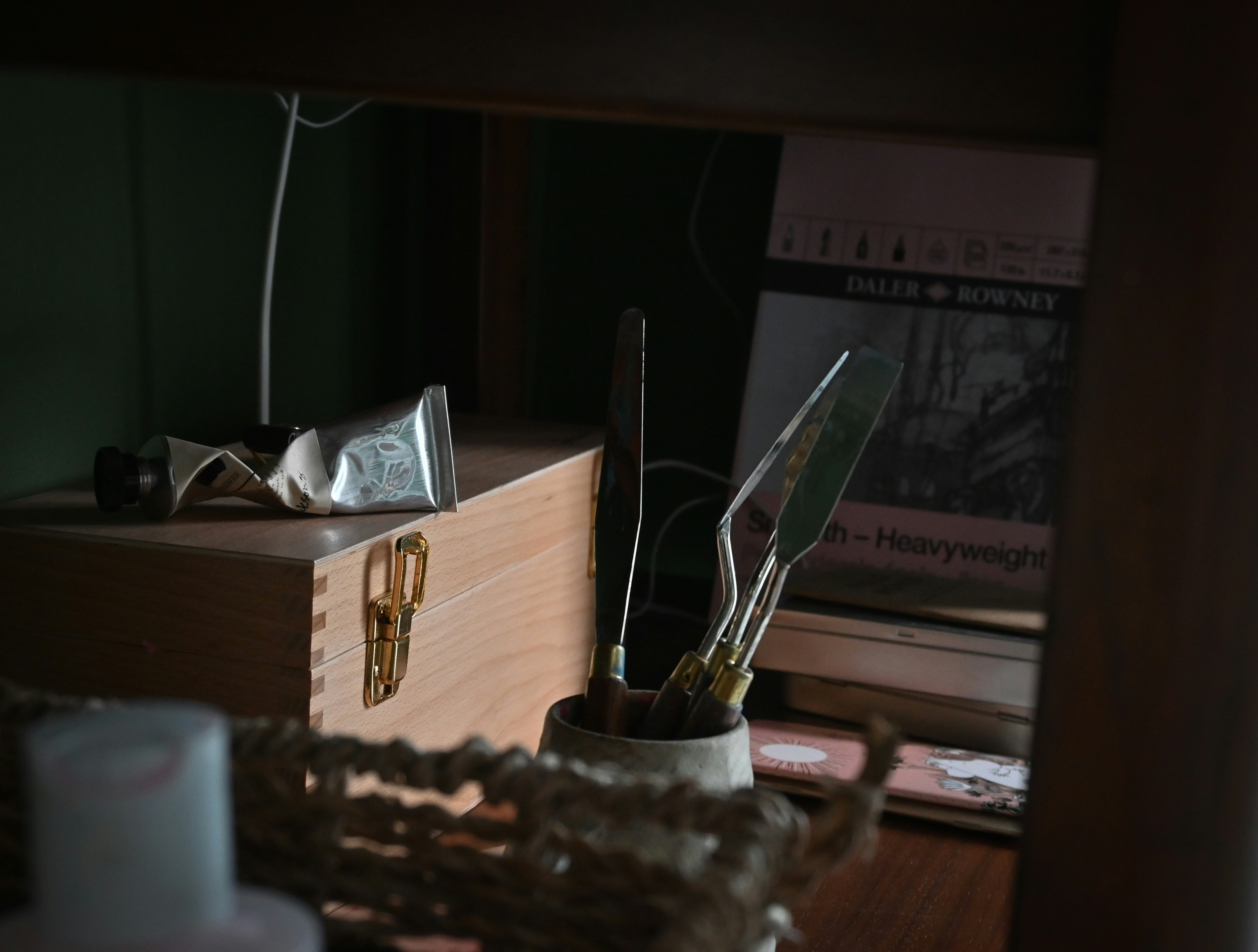 A wooden box and various artist tools, including knives and a tube of paint, arranged on a shelf with a hint of vintage decor.