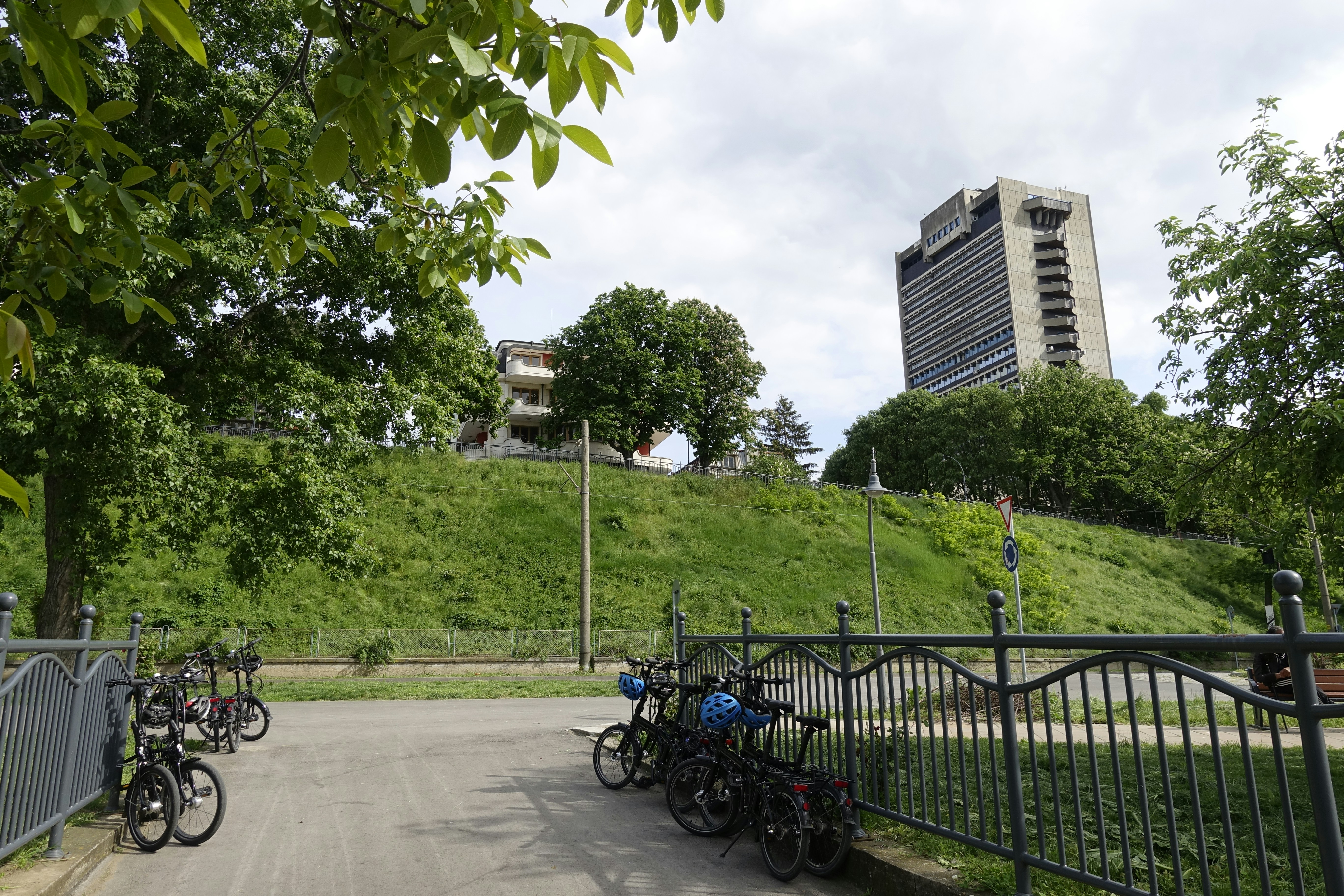 Bicycles parked along a path with a grassy hill and tall building in the background.