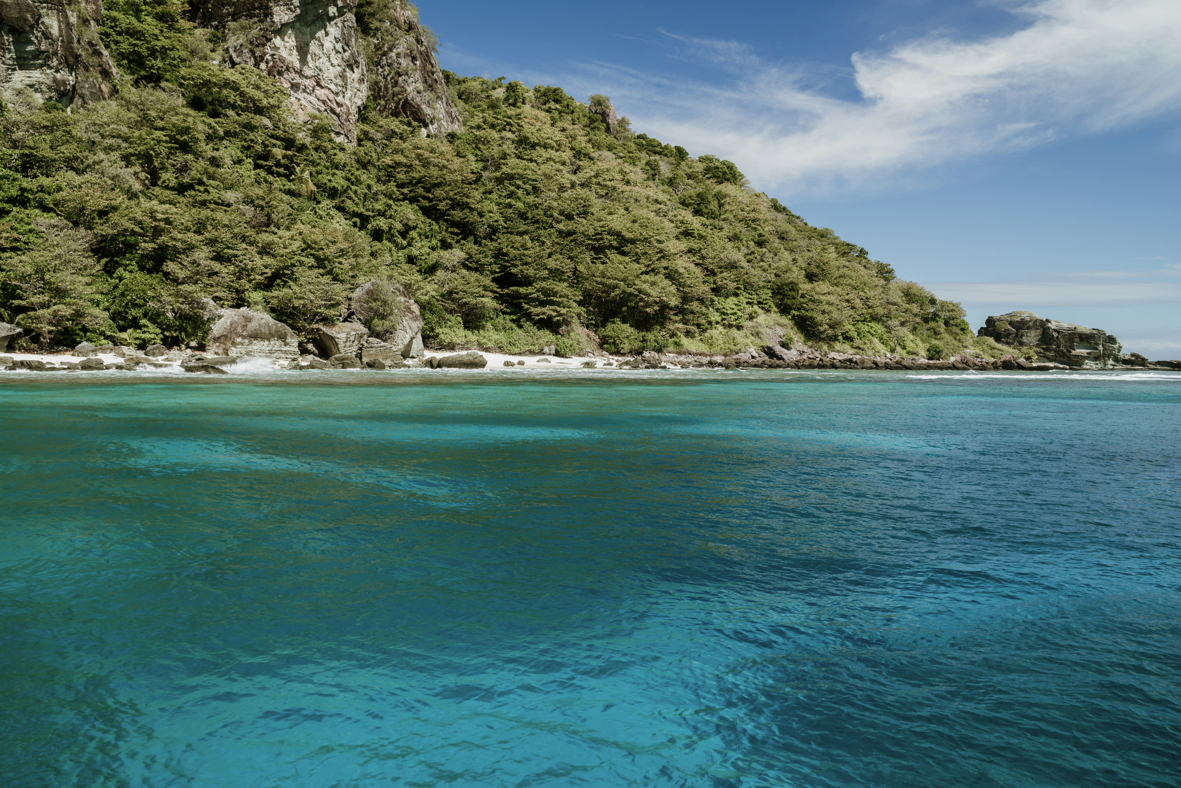 This captivating image showcases a lush, green hillside meeting the tranquil turquoise waters of the ocean, under a bright blue sky with wispy clouds. The vibrant color palette of deep blues and greens contrasts beautifully, creating a serene and inviting atmosphere. The composition draws the eye from the foreground water to the rich textures of the foliage, highlighting the natural beauty of this coastal paradise.