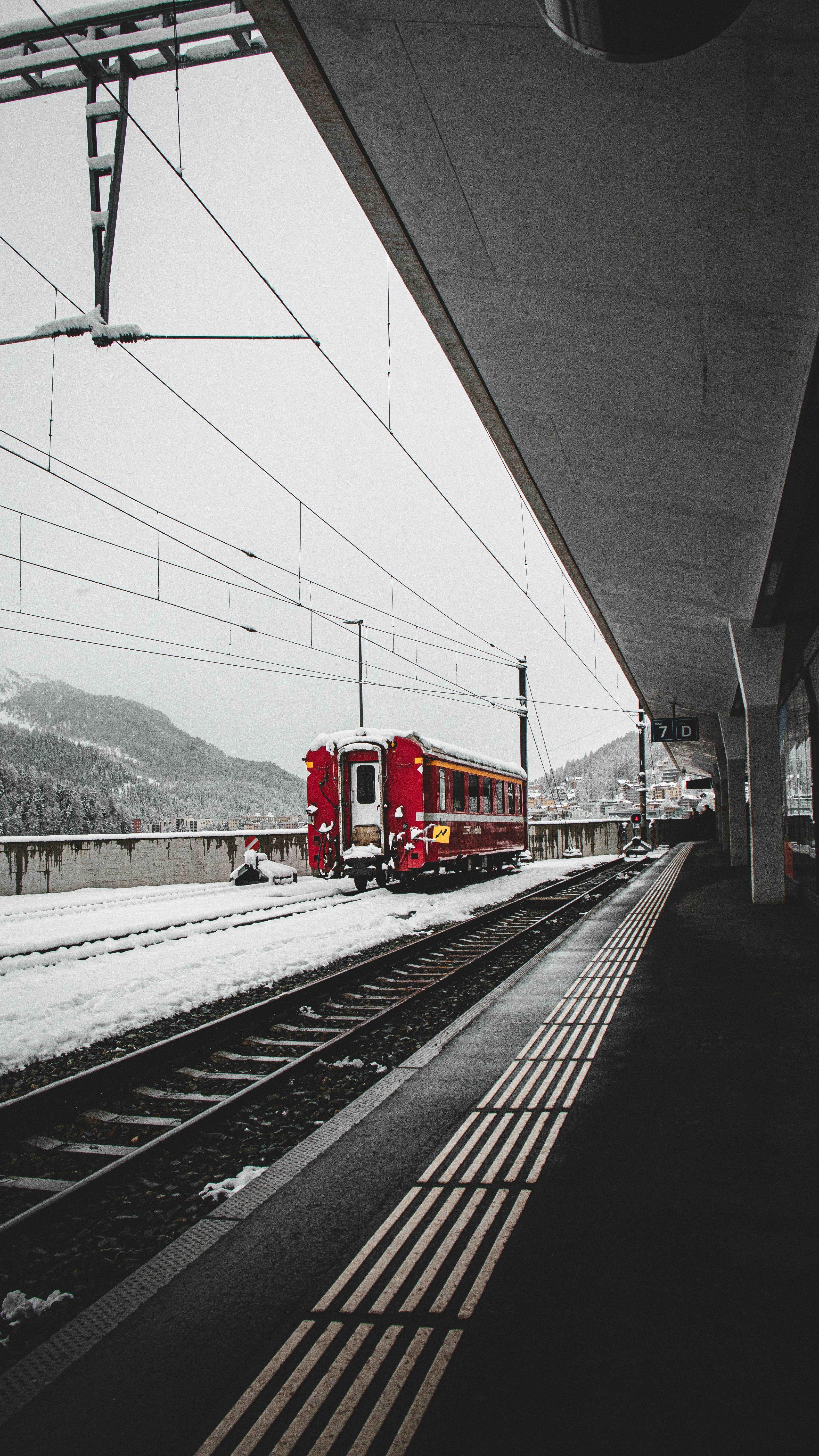A vibrant red train sits on snowy tracks at a mountain station, surrounded by a serene winter landscape. The scene captures the essence of travel in a snowy environment.