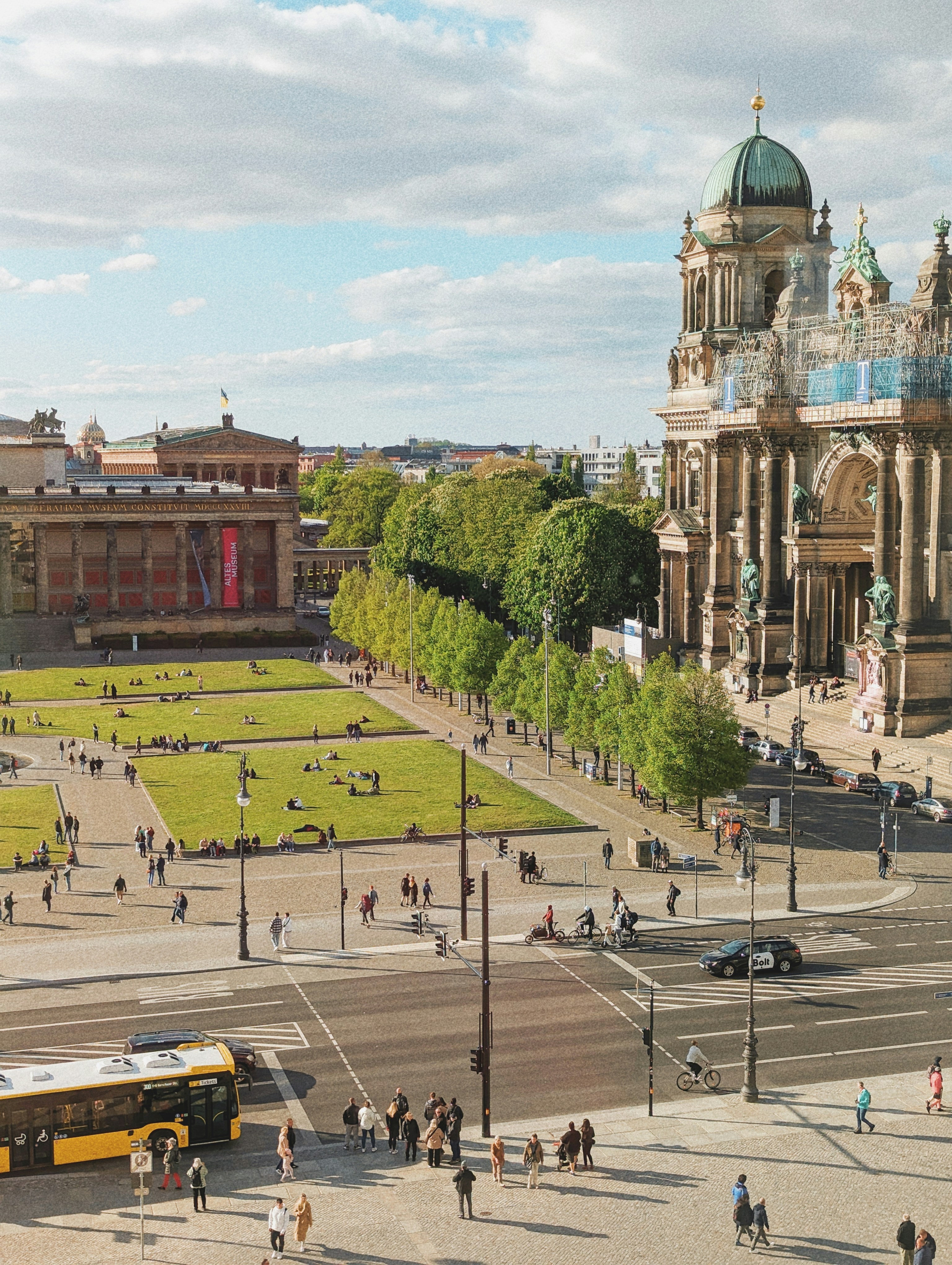 Wide-angle street-level view shows a grand domed cathedral on the right, a tree-lined square, and pedestrians with a yellow bus at the curb.