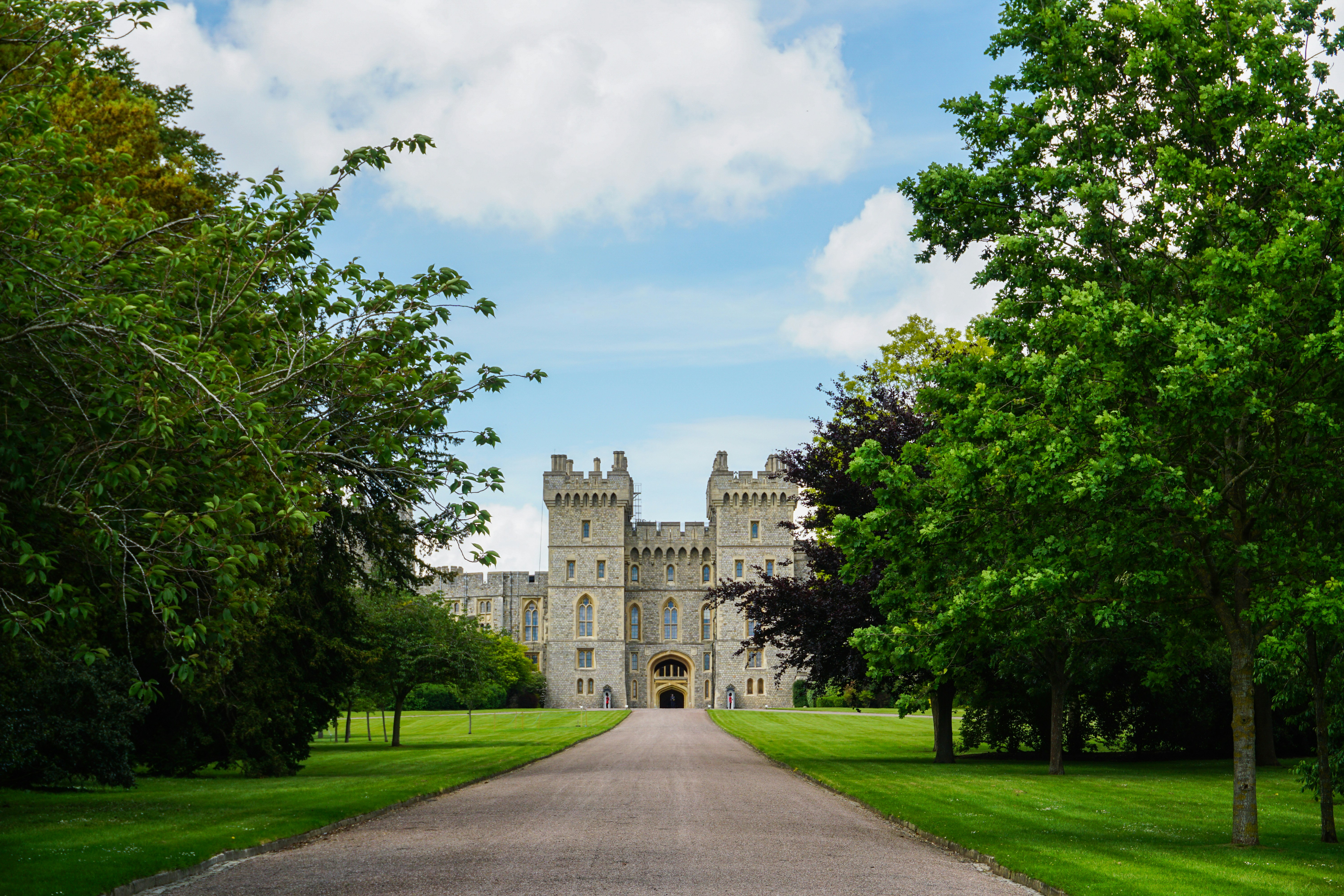 Long driveway flanked by lush trees leading to a large castle under a partly cloudy sky.