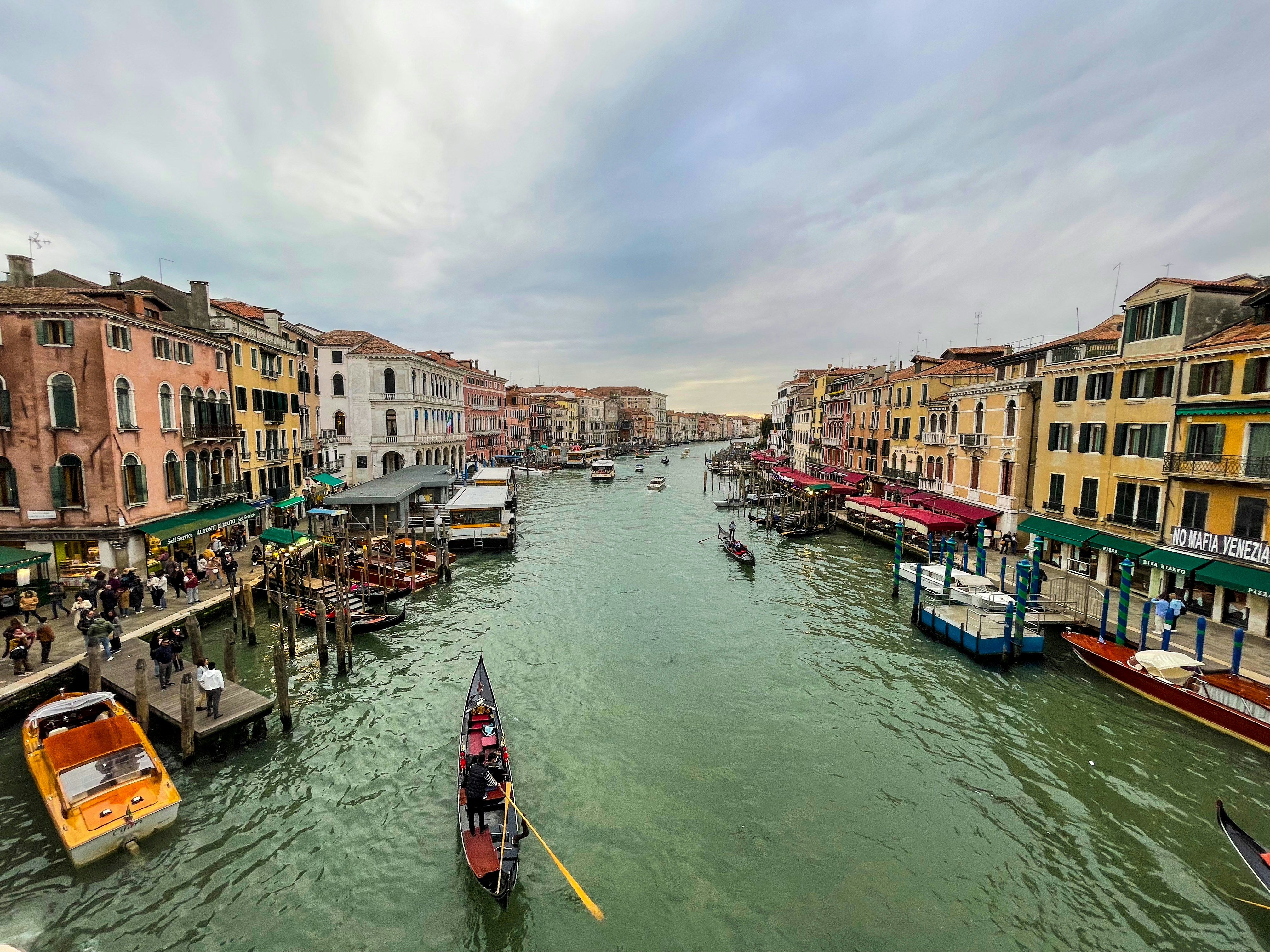 Gondolas glide along a bustling canal flanked by historic Venetian buildings under a cloudy sky.