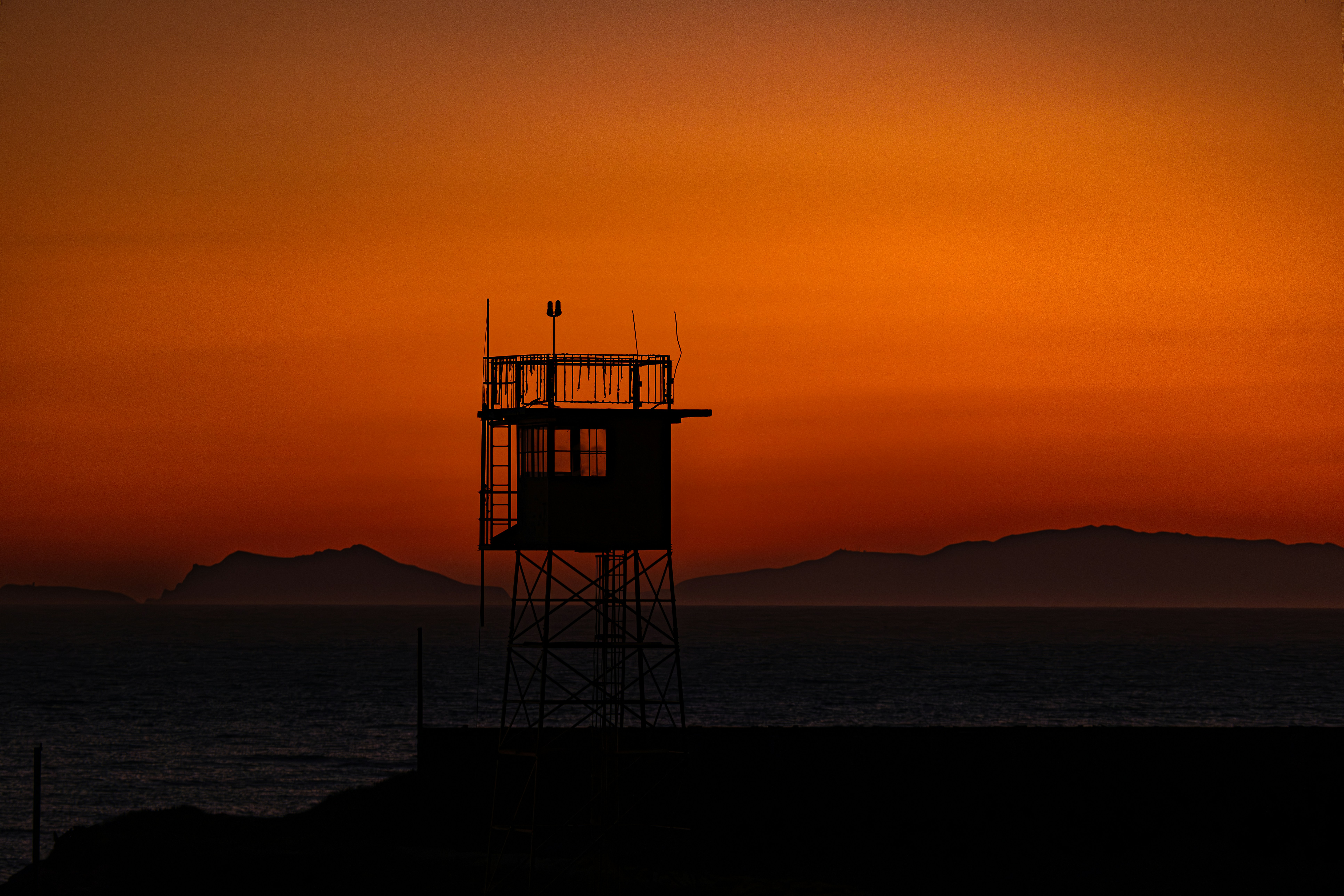 Silhouette of a metal watchtower on a headland against a fiery sunset over the sea, with distant mountains on the horizon.