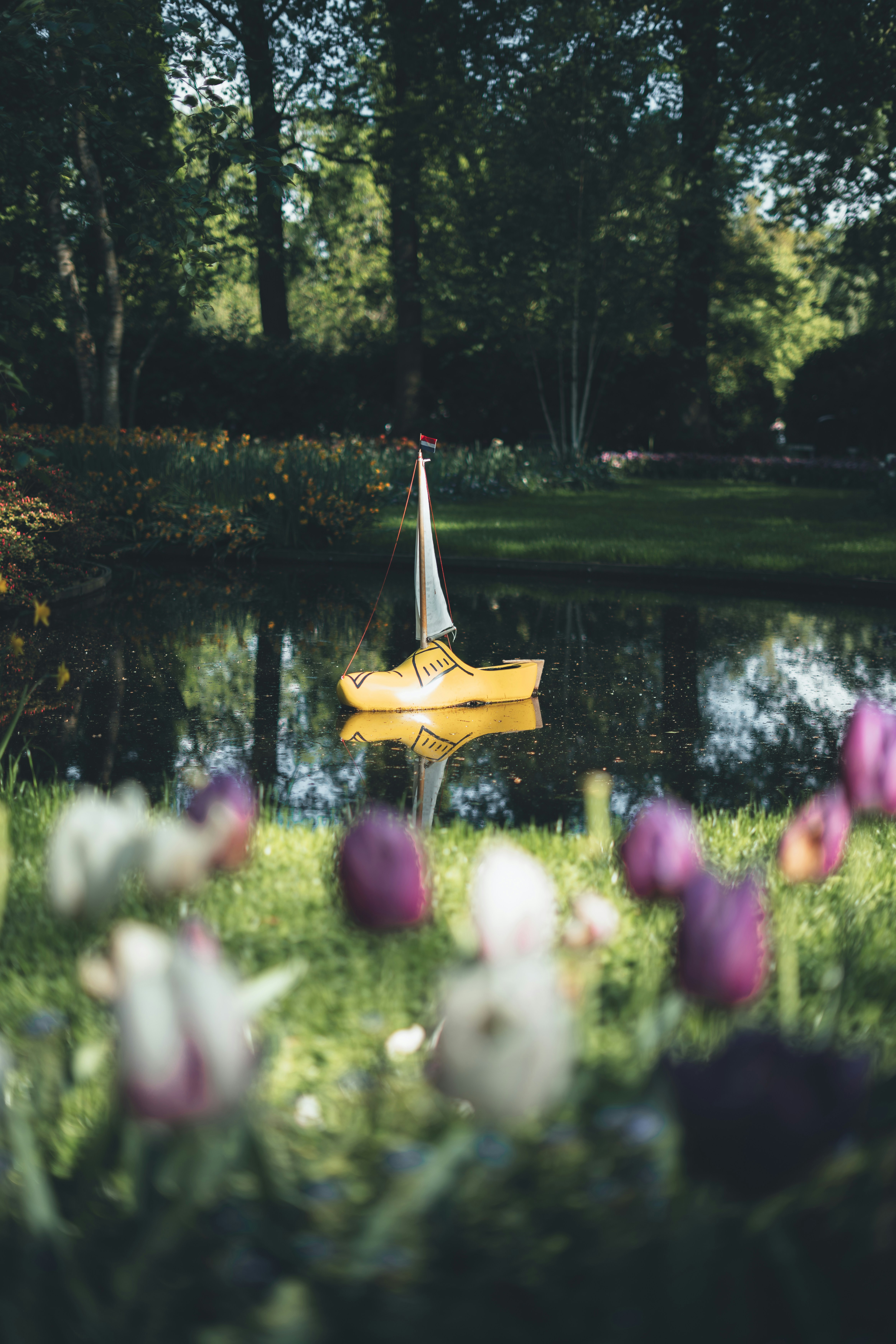 Yellow toy sailboat on a tranquil pond, mirrored by the water and framed by blurred tulips