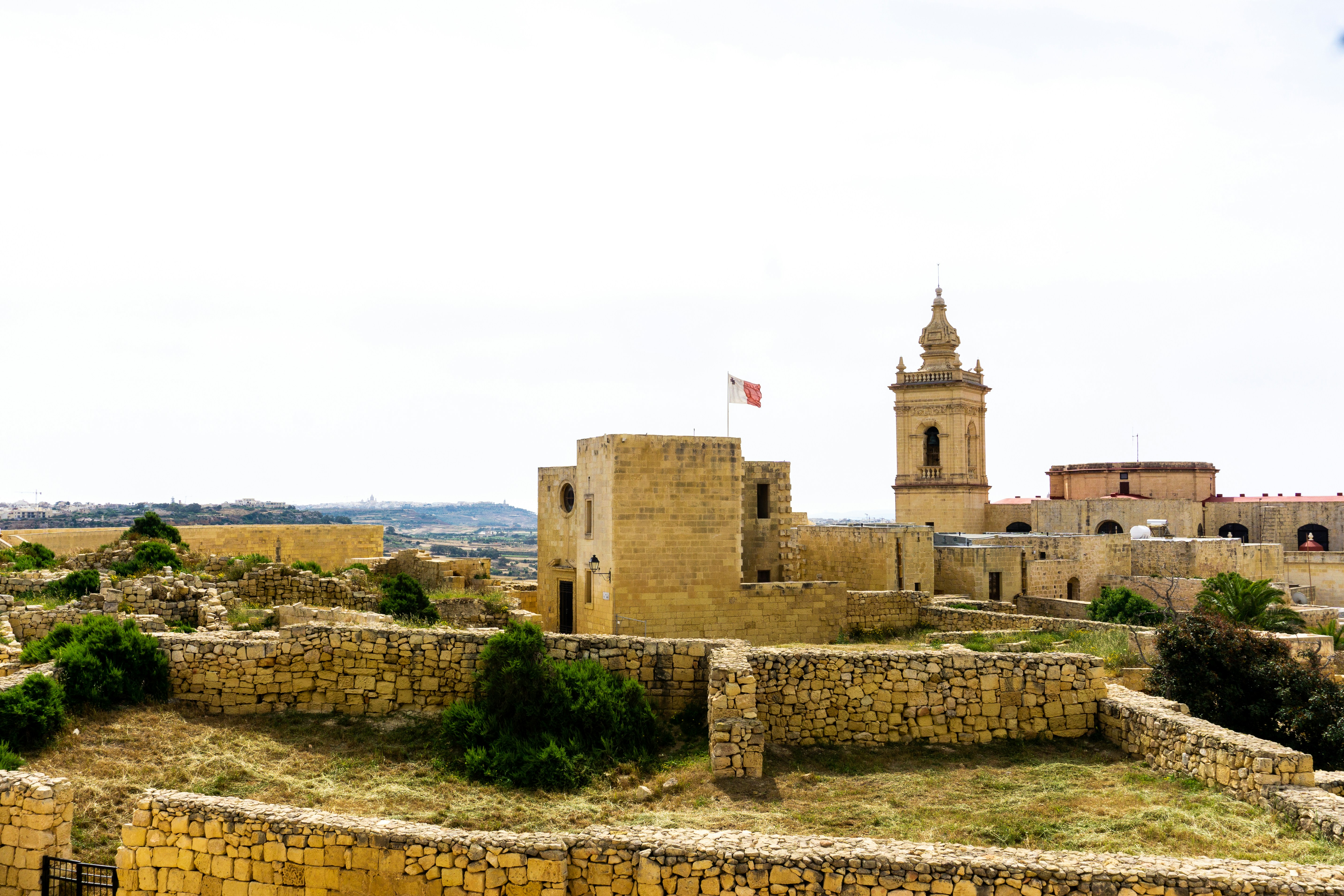 Stone castle with clock tower surrounded by ancient walls and greenery under a bright sky.