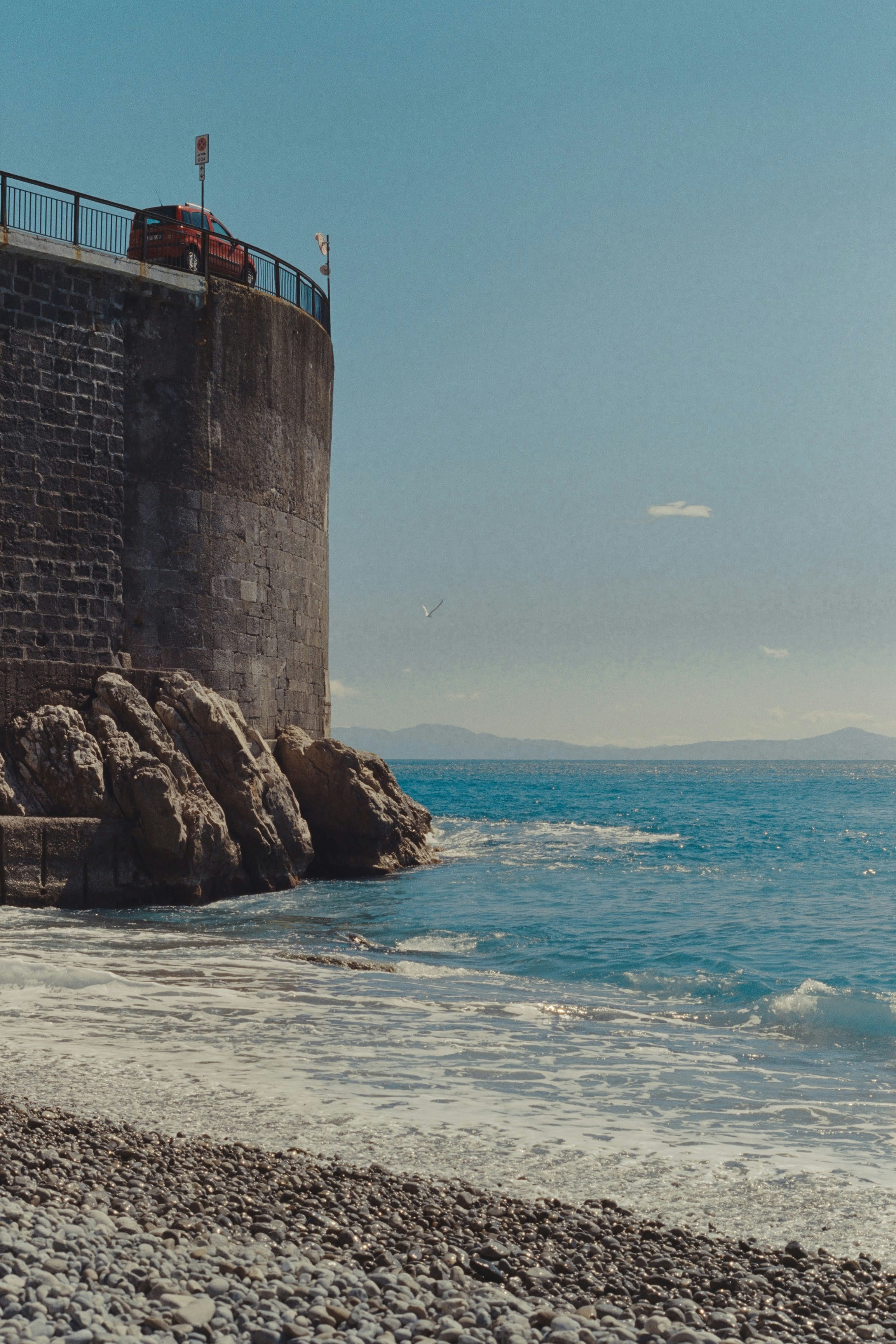 Coastal Sentinel: A Red Car Guards the Stone Citadel Over Calm Seas