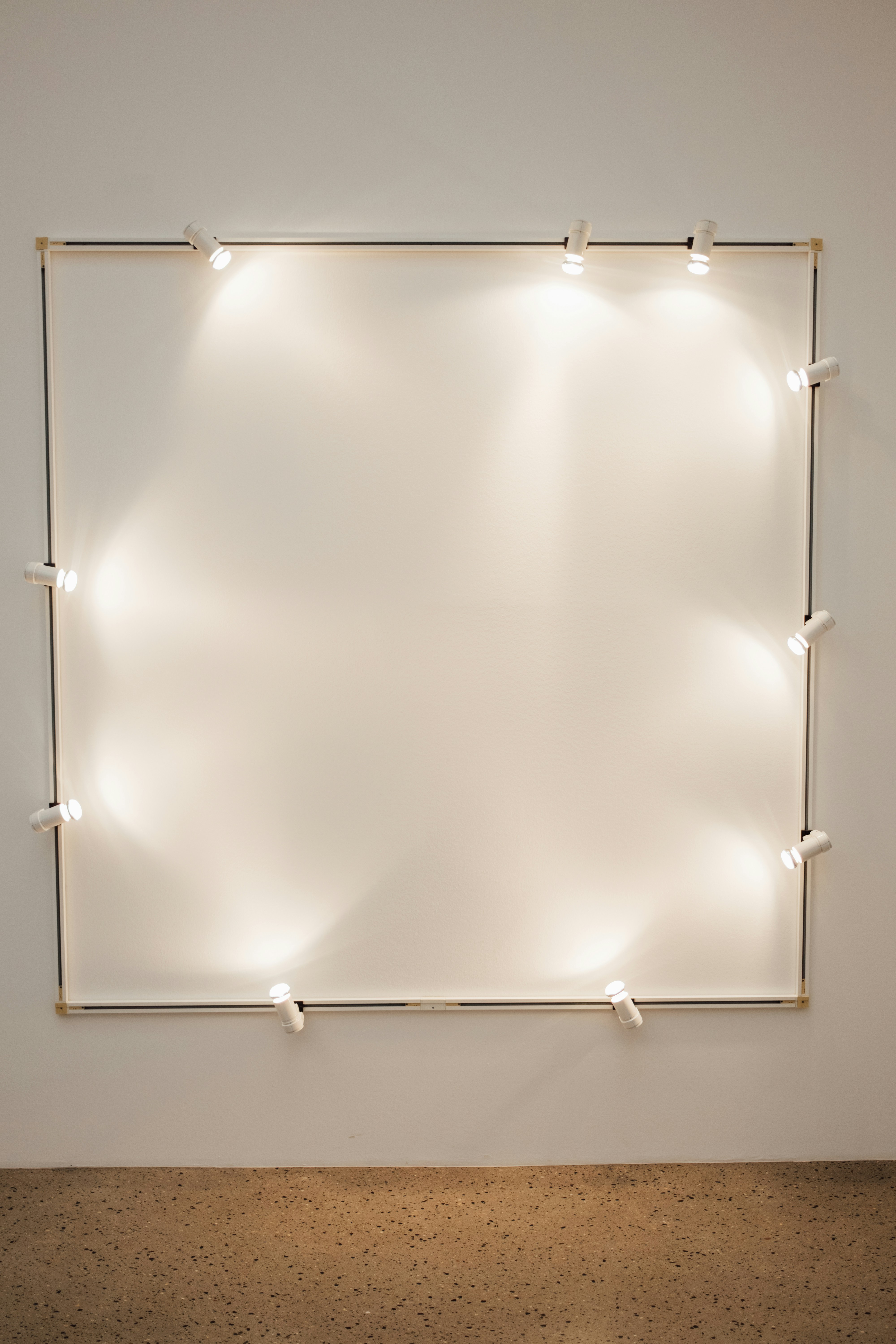 Softly illuminated square frame with exposed bulbs mounted to a white wall, resting against a neutral terrazzo floor. A clean, minimal setup highlighting a studio lighting concept.