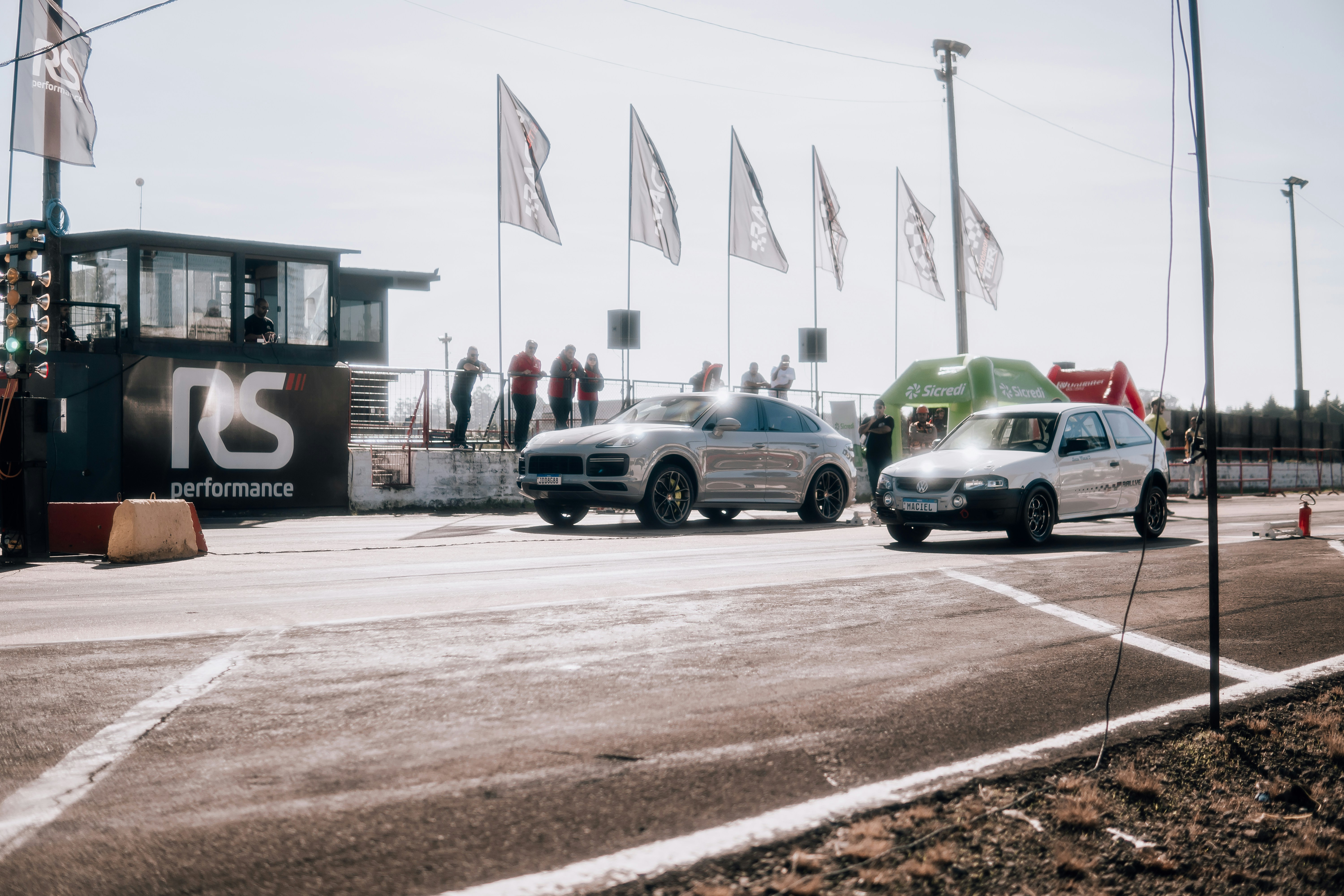 Two cars positioned at the starting line of a racetrack with spectators and flags in the background.
