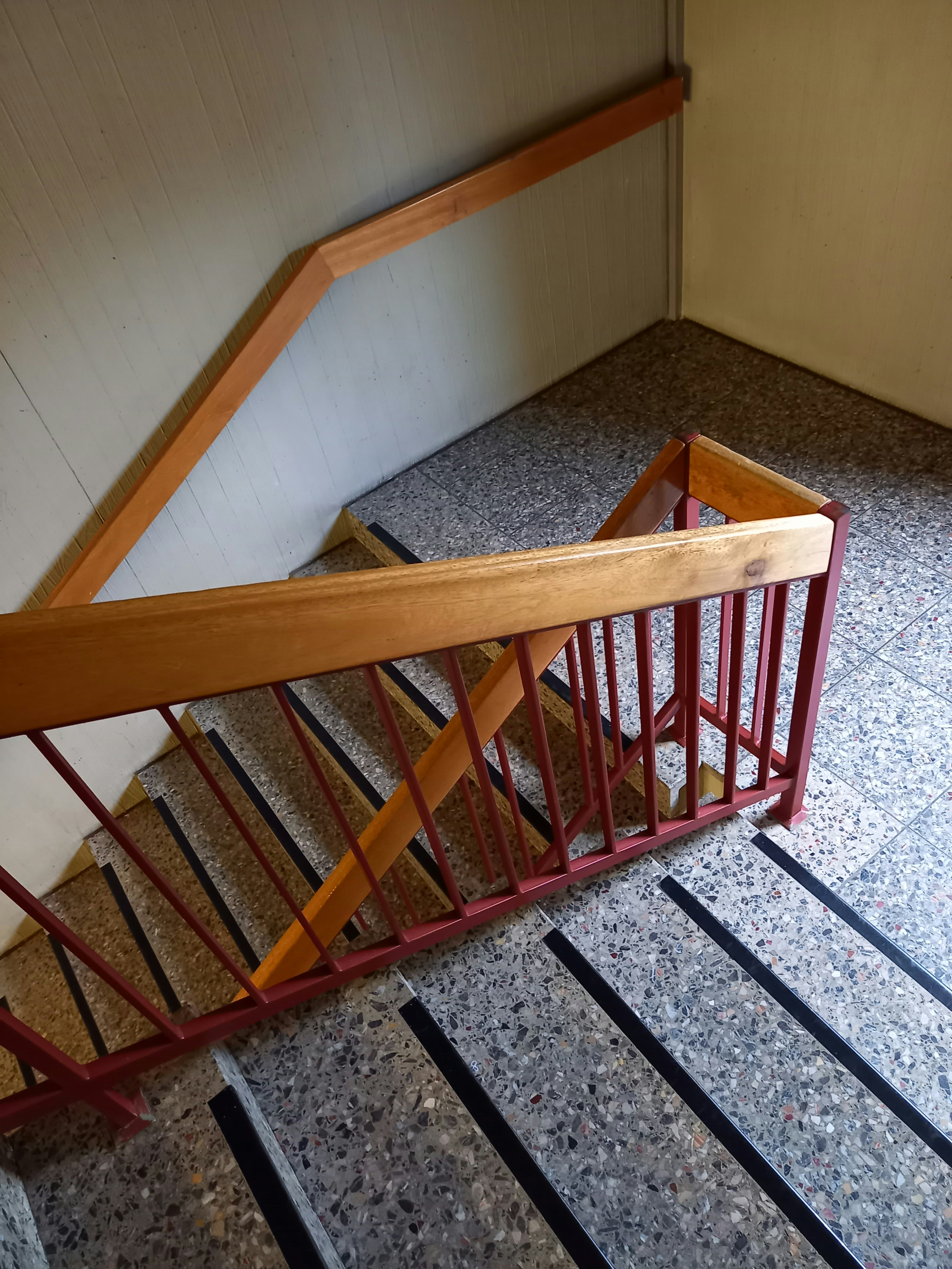 A tilted view of a wooden handrail and red metal balusters tracing a staircase toward a corner; terrazzo steps and pale walls form a geometric, converging perspective.