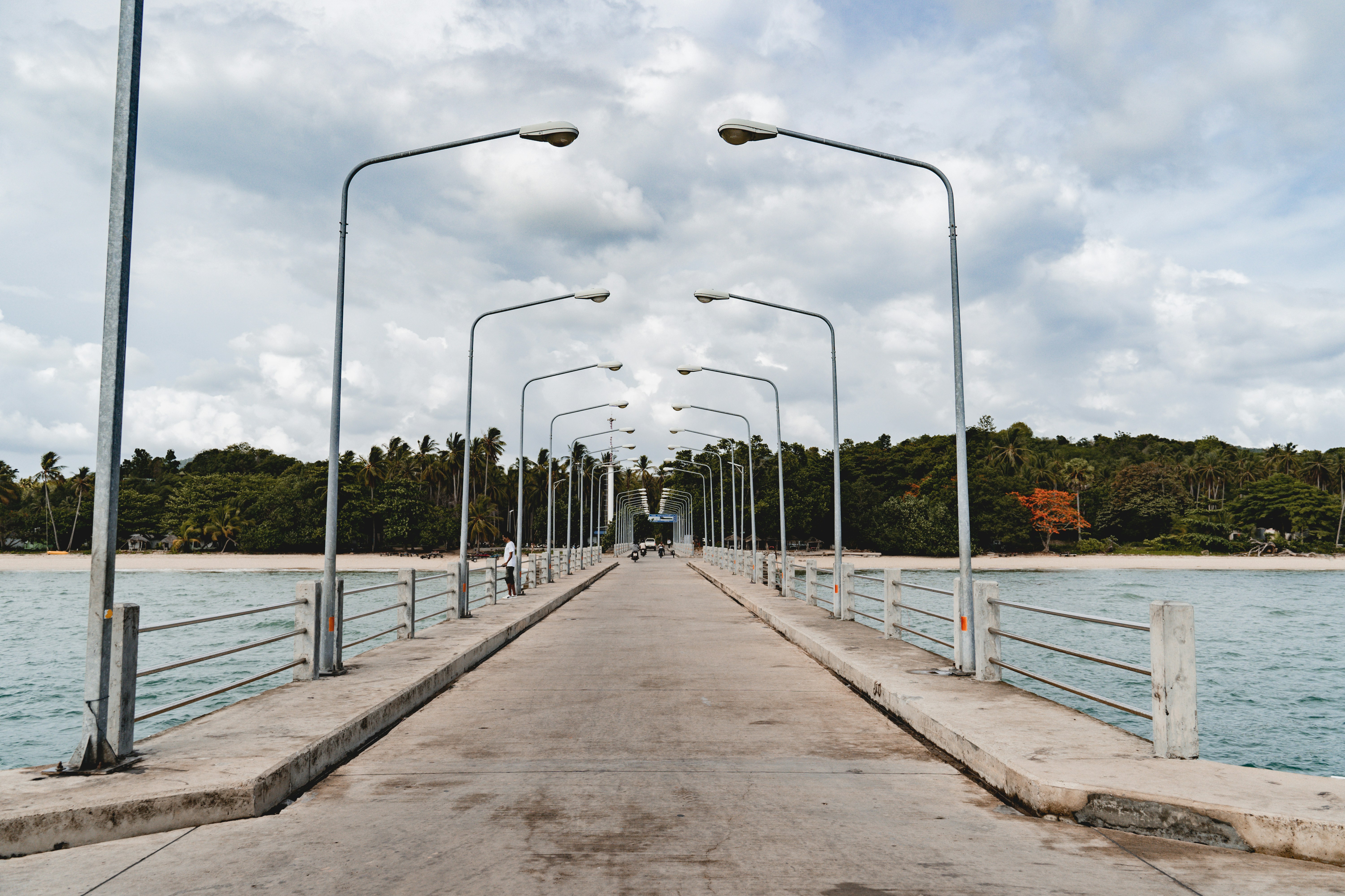 Concrete pier lined with streetlights extending towards a distant shoreline under a cloudy sky.