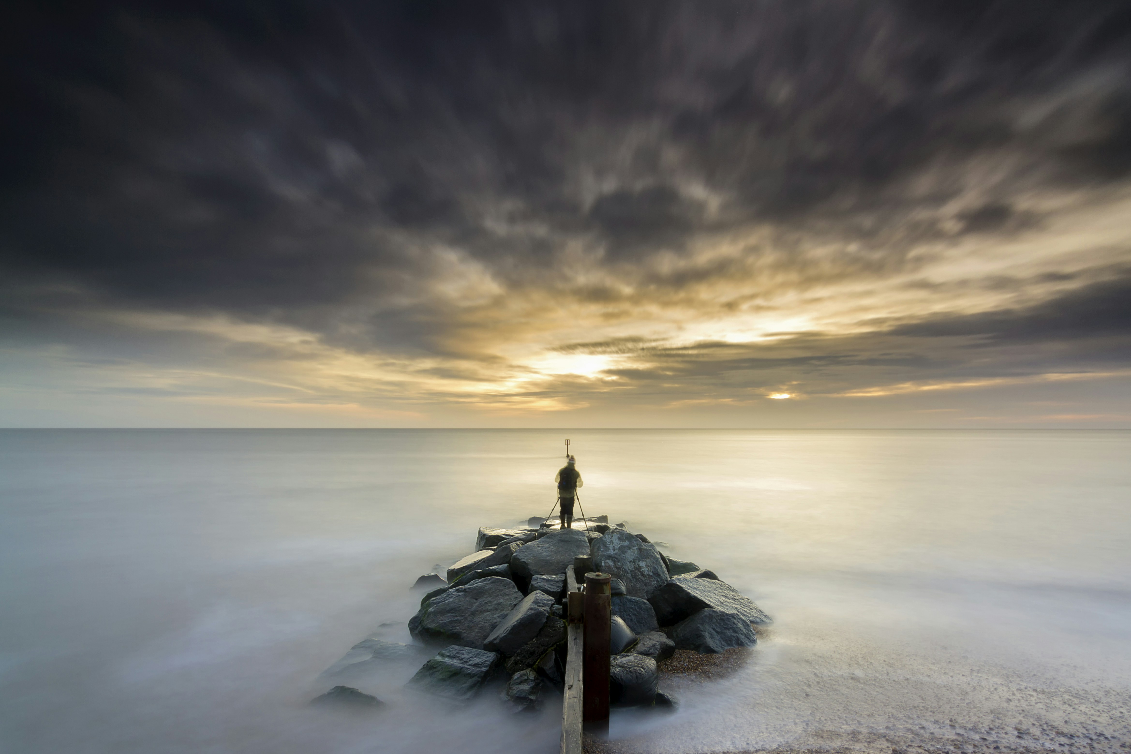 A lone figure stands on a rocky jetty, gazing out over a serene ocean at sunset, with dramatic clouds swirling overhead.