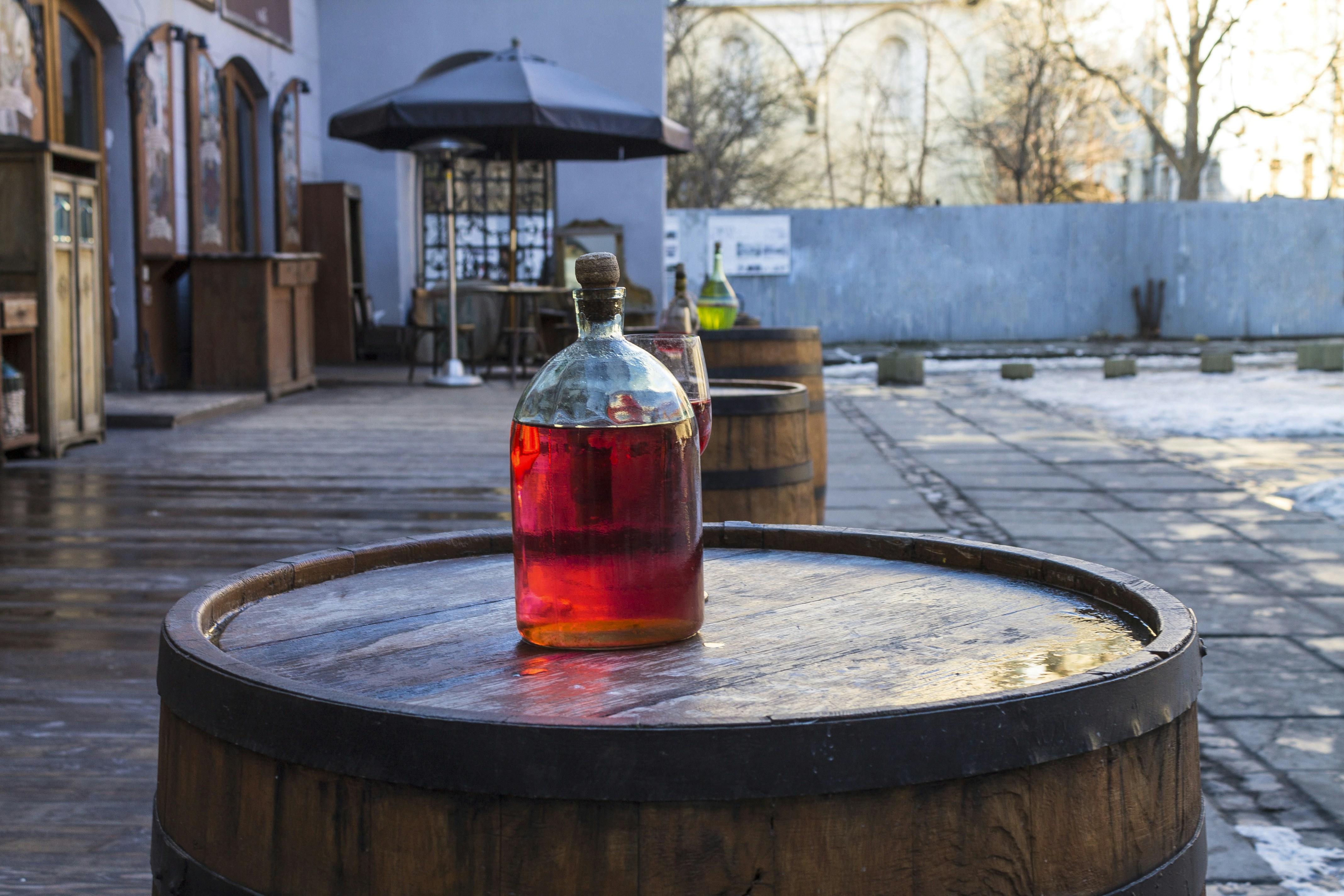 Glass bottle filled with a colorful liquid sits on a wooden barrel, surrounded by an outdoor rustic atmosphere. The scene captures the essence of a cozy gathering space.