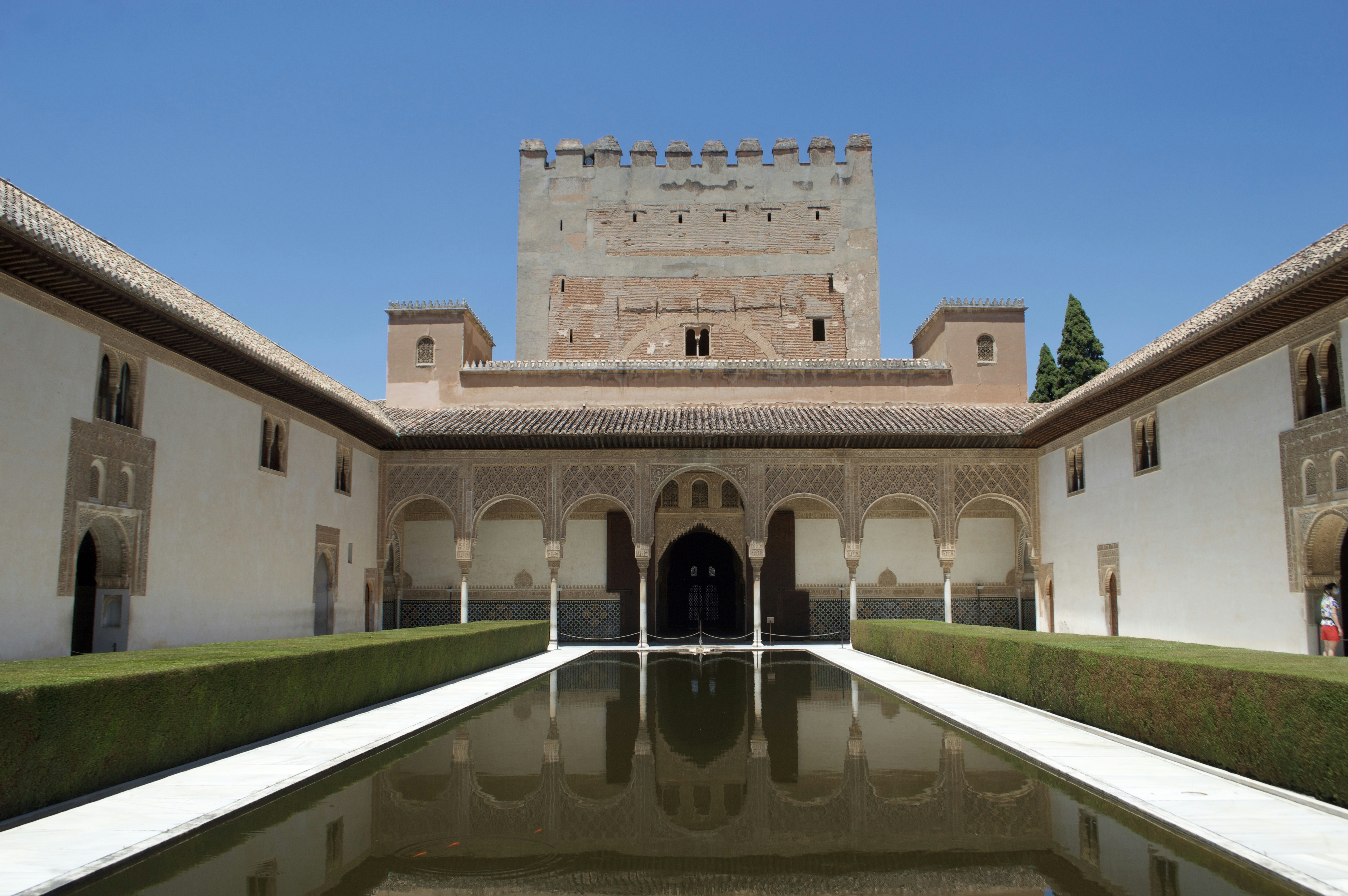 Symmetrical courtyard with arched openings reflected in a central pool under a clear blue sky.