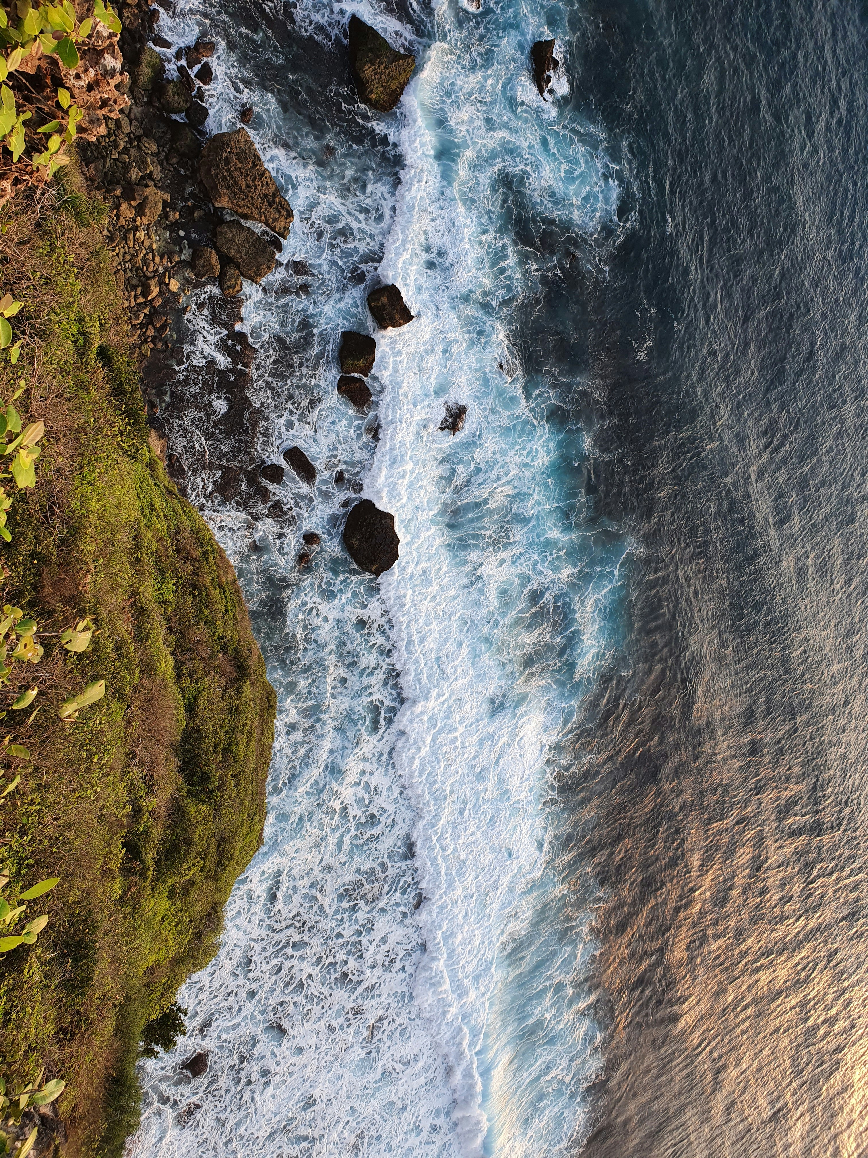 Waves crashing against rocky cliffs with lush greenery at sunset.