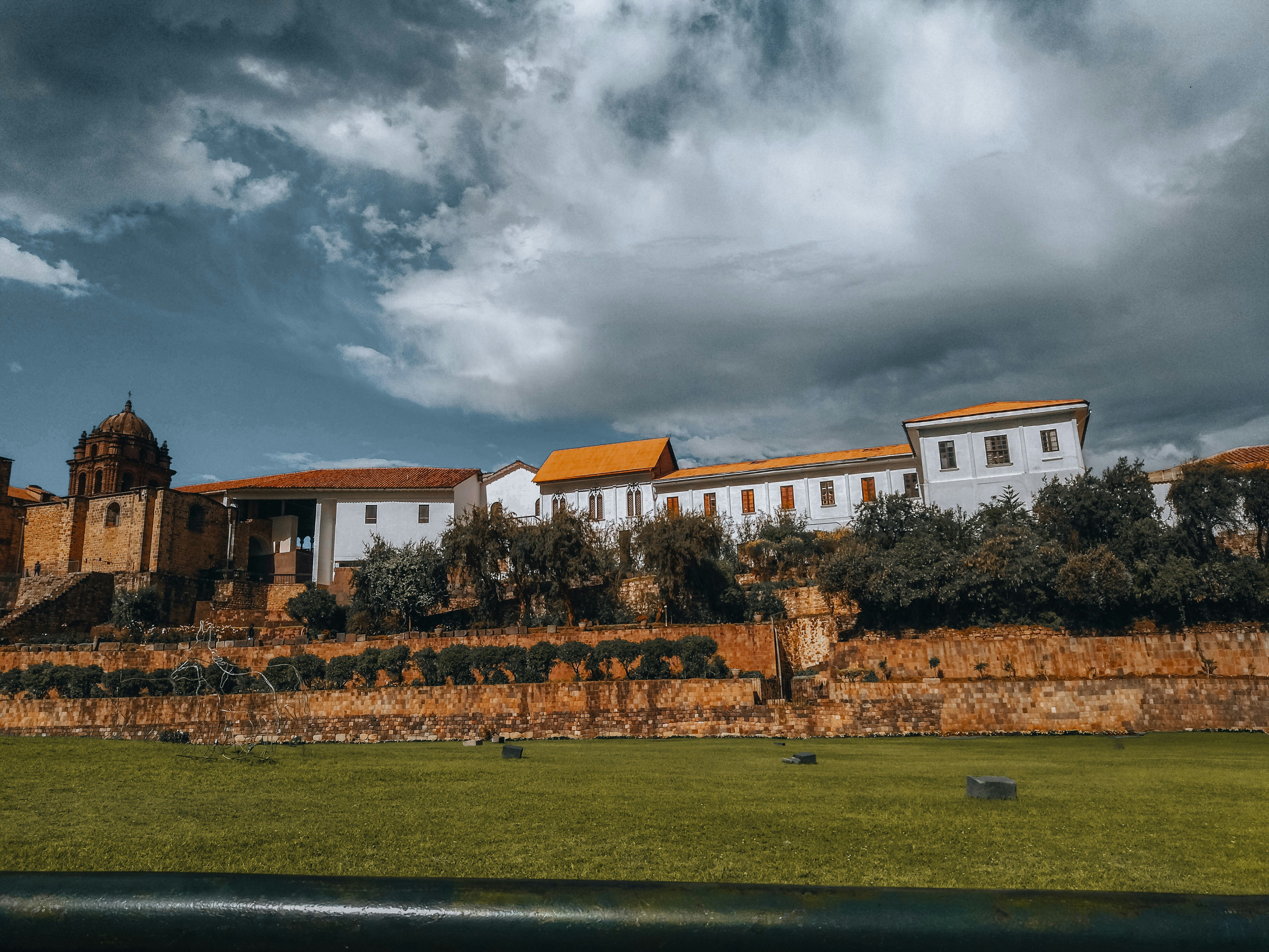 Large white building with terracotta roofs perched on a lush green field under a dramatic sky.