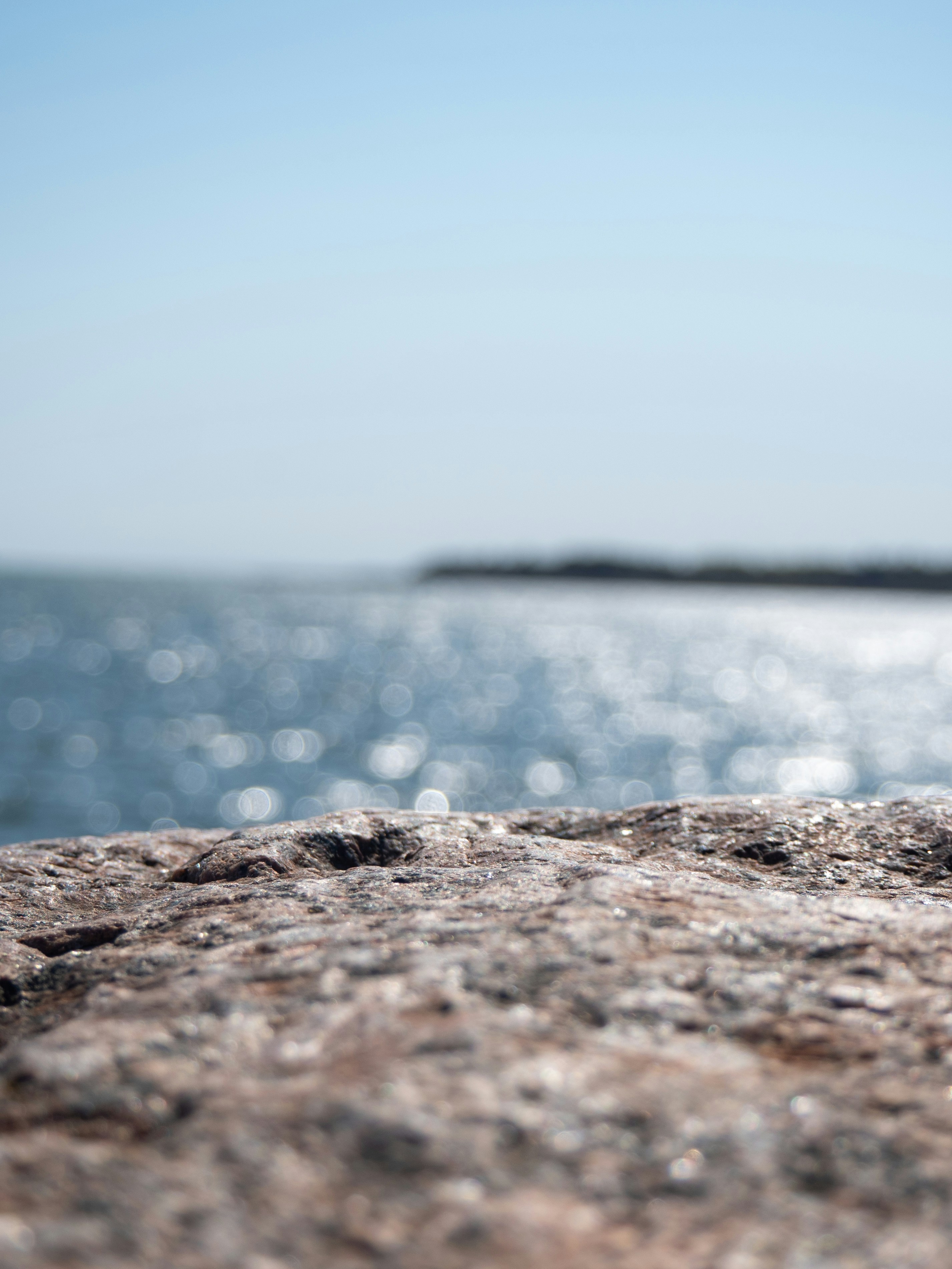 A shallow-depth photograph capturing textured rock in the foreground with a blurred, sparkling ocean and distant horizon.