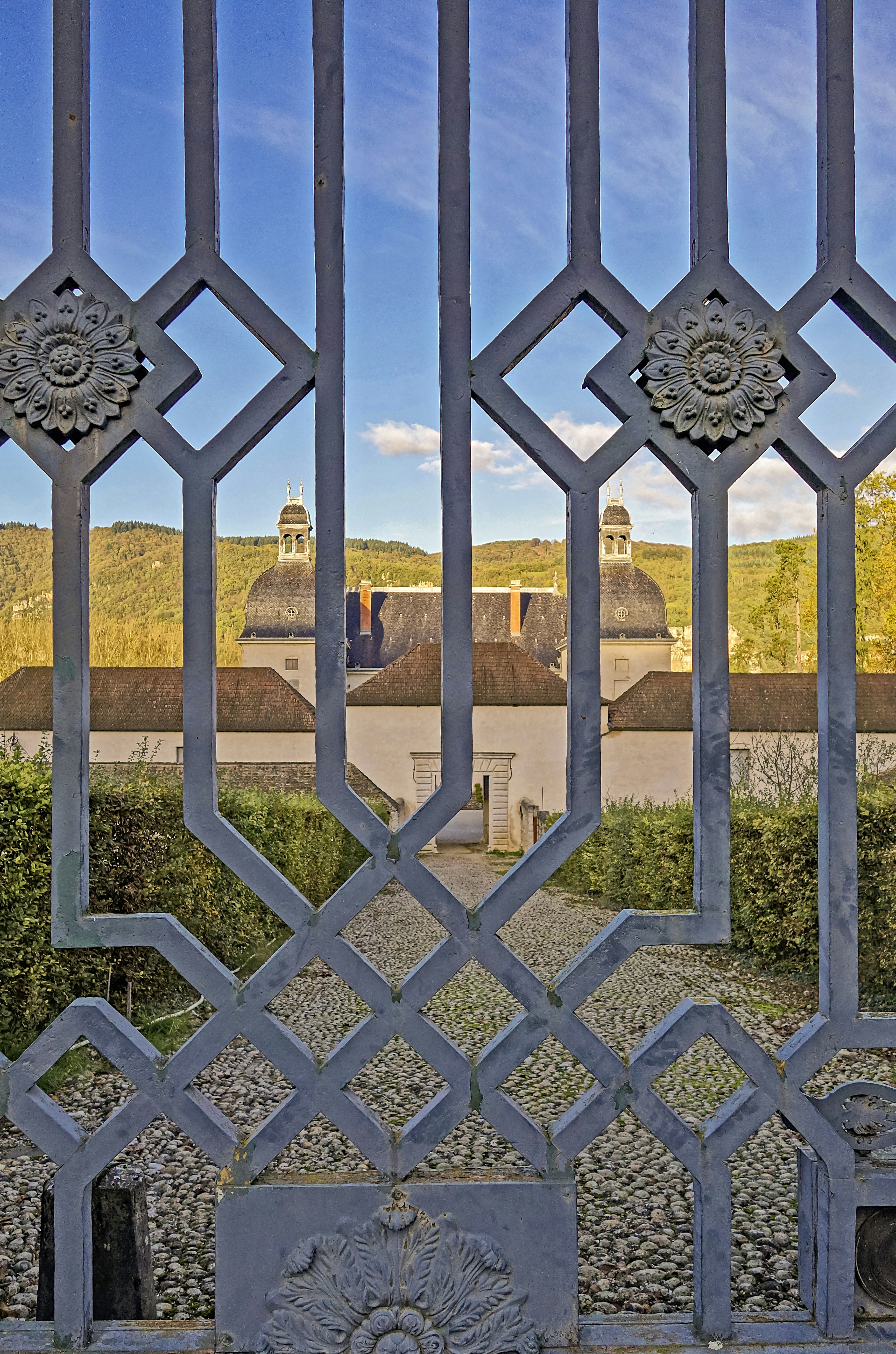 Ornate metal gate framing a historic building with domed towers against a backdrop of rolling hills.