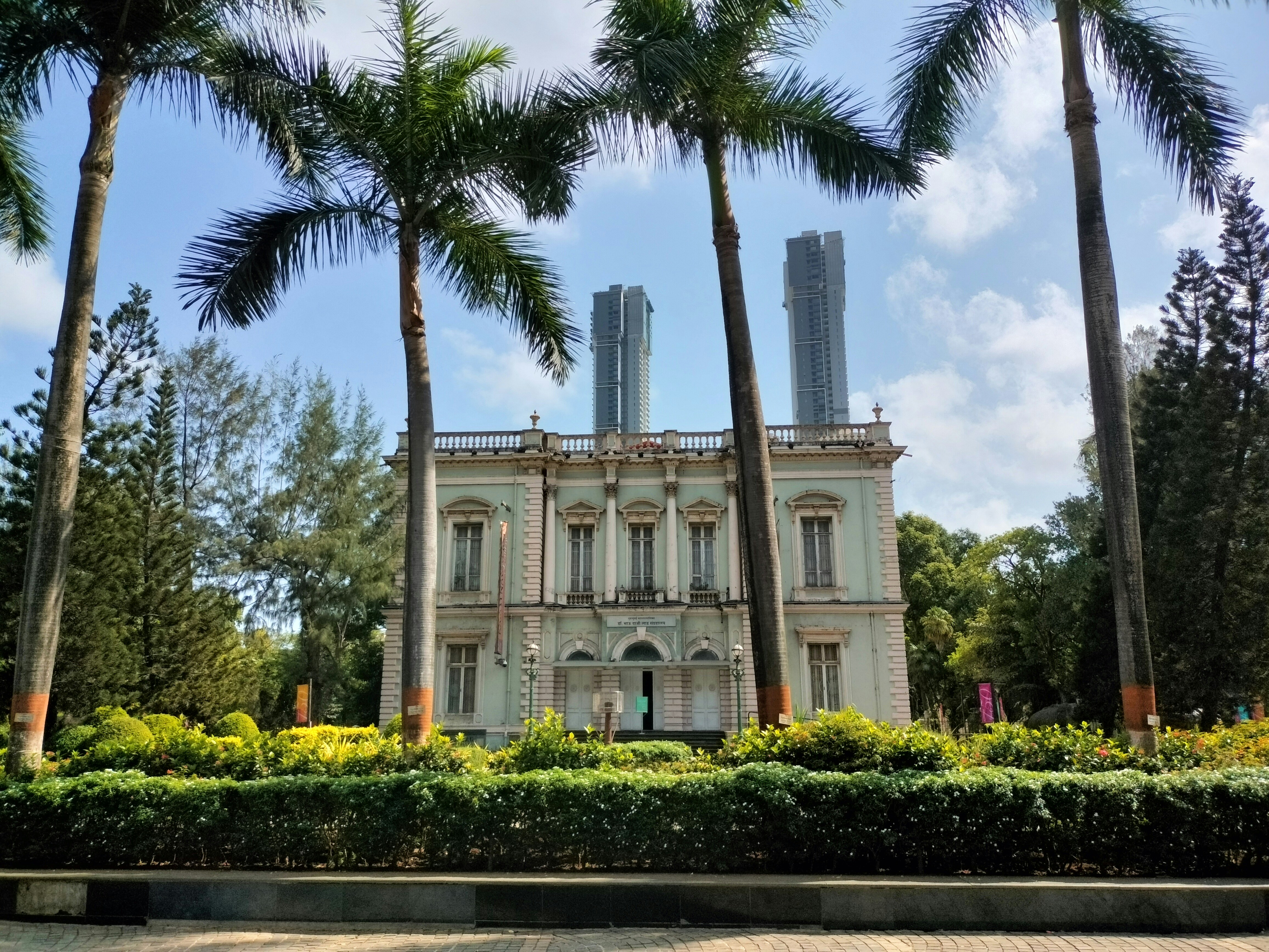 A neoclassical building sits in a manicured garden framed by tall palm trees. Modern skyscrapers rise in the distance, creating a layered city backdrop.