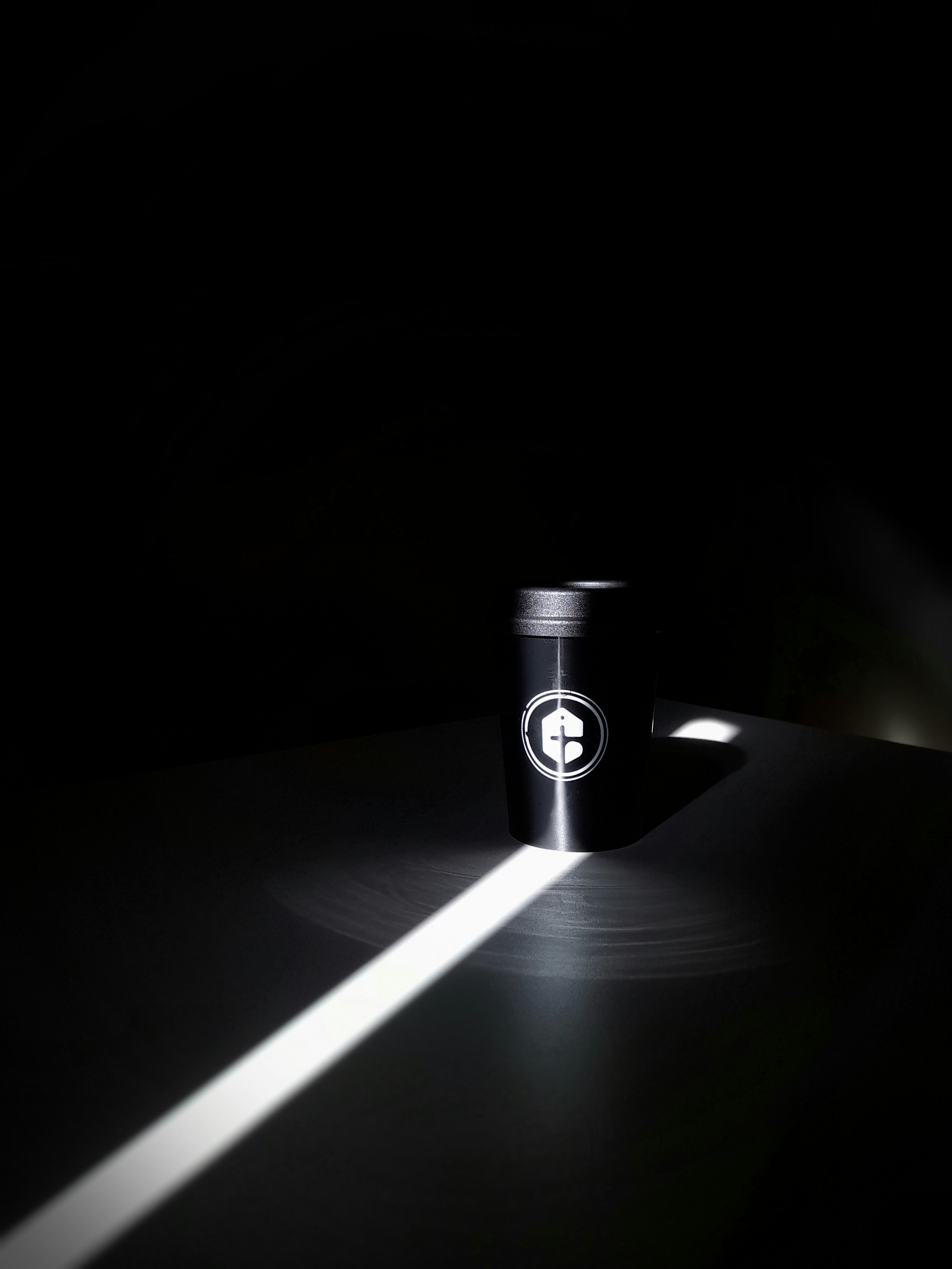 Low-key shot of a cylindrical object with a logo on a dark table, illuminated by a single beam of light creating a glossy highlight and circular reflections on the surface.