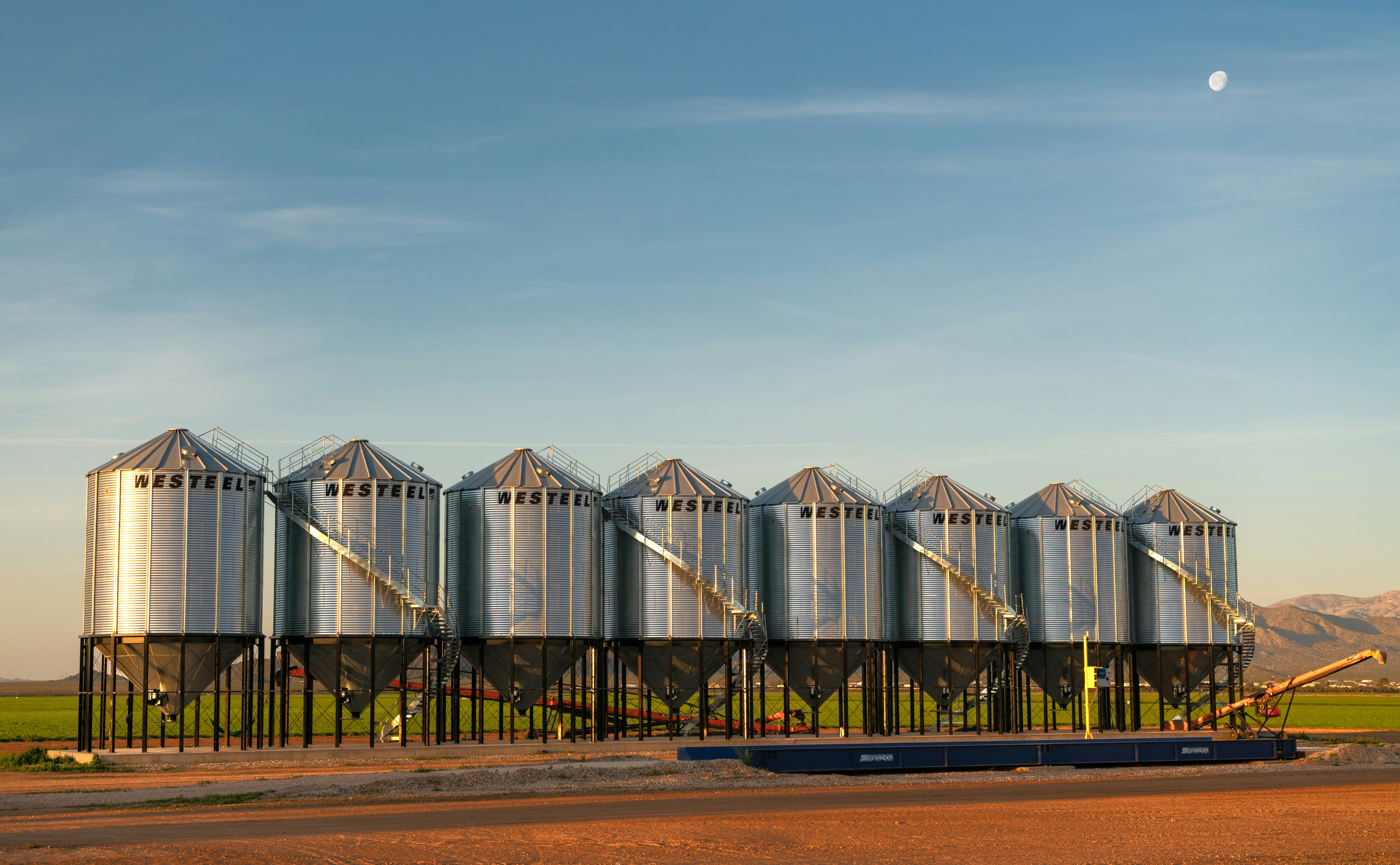 Row of metallic grain silos under a clear sky with a rising moon, showcasing agricultural infrastructure.