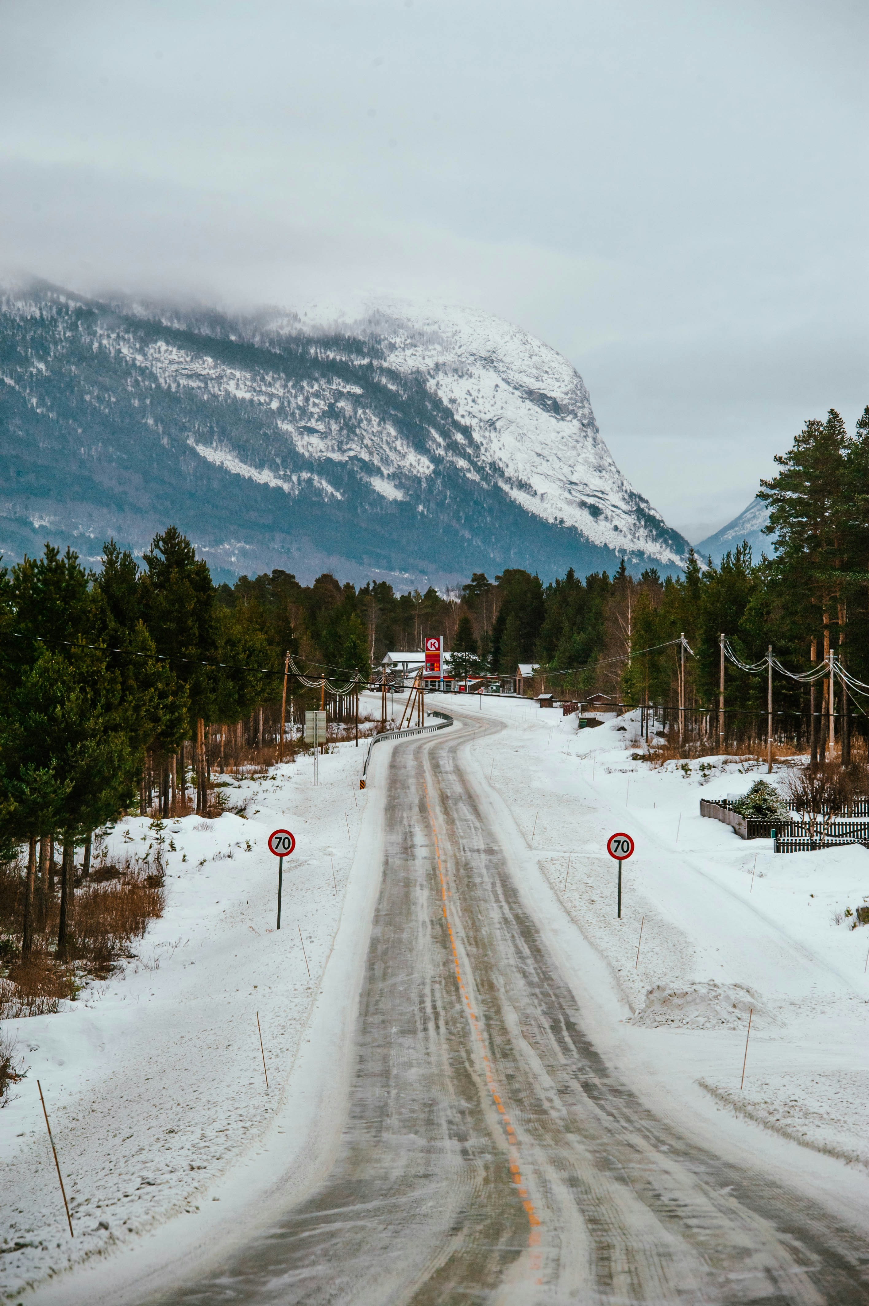 Snow-covered road winds toward a snow-capped mountain, framed by evergreen trees under a gray sky.