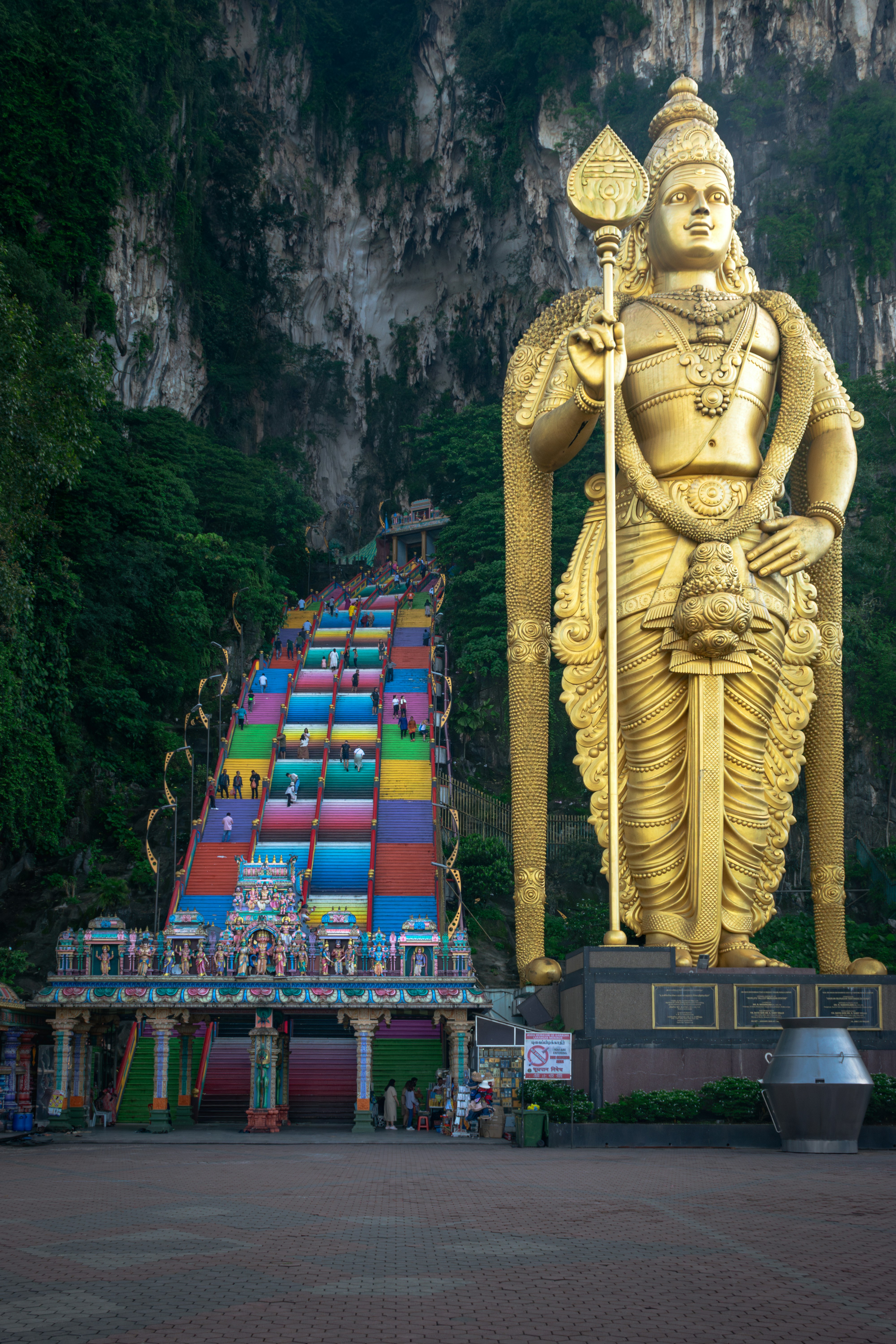 Majestic golden statue of a deity stands guard over the vibrant steps leading to Batu Caves, surrounded by lush greenery.