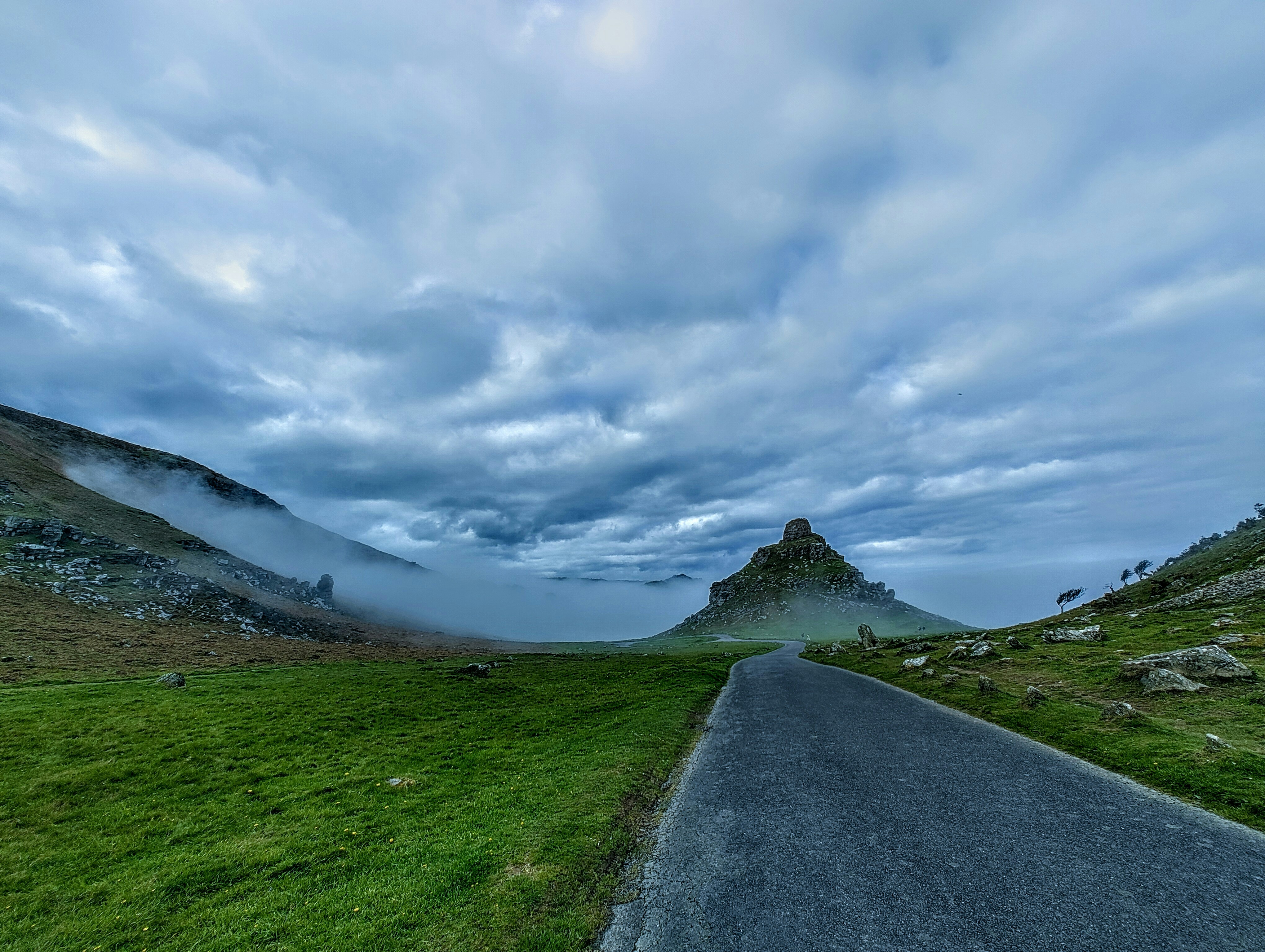 Landscape photograph of a paved road winding through green hills toward a solitary rock peak, with fog drifting over the valley and a dramatic, cloud-filled sky.